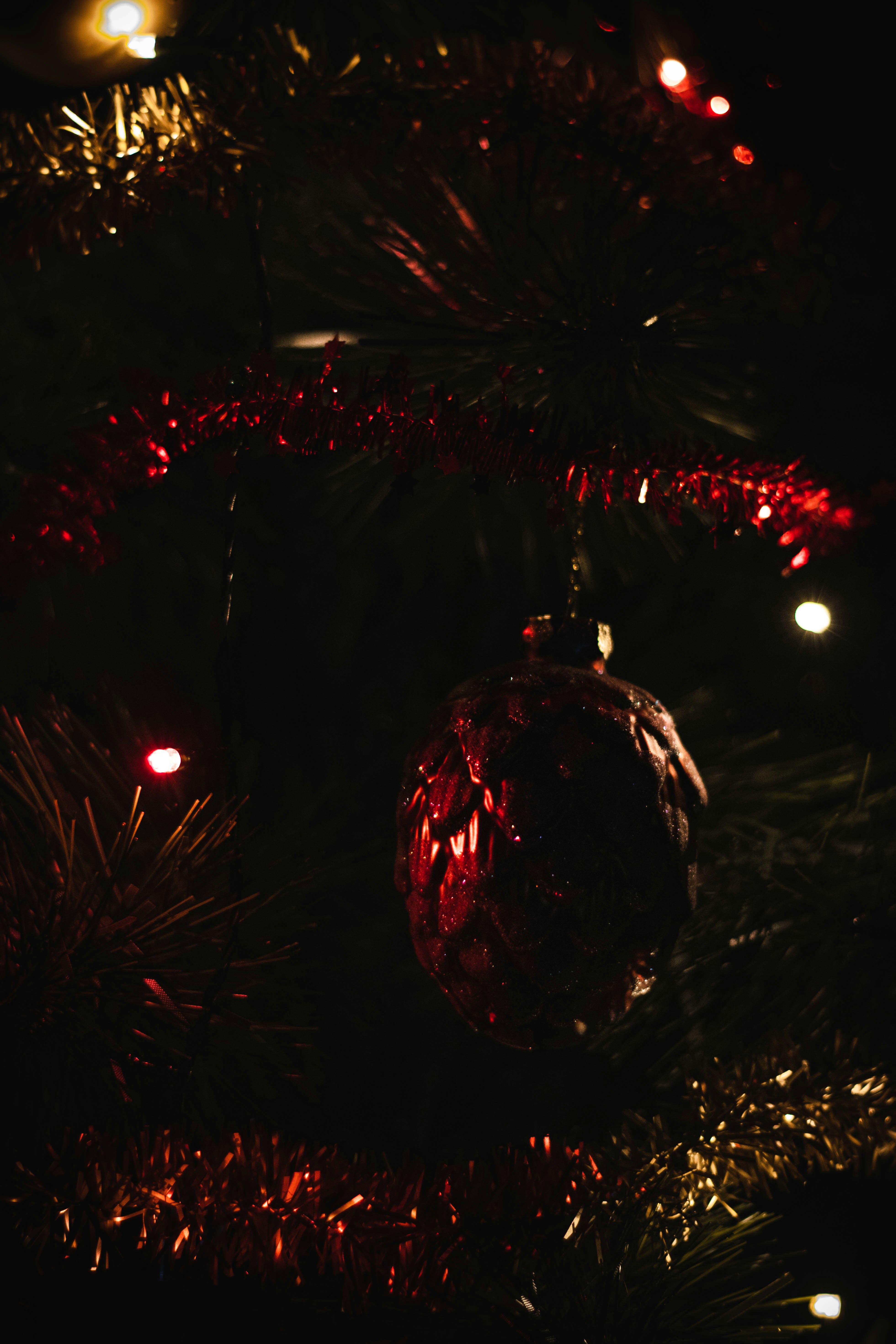 A shimmering pine cone ornament hangs delicately from a Christmas tree, surrounded by softly glowing lights and tinsel. The dark backdrop enhances the festive atmosphere.