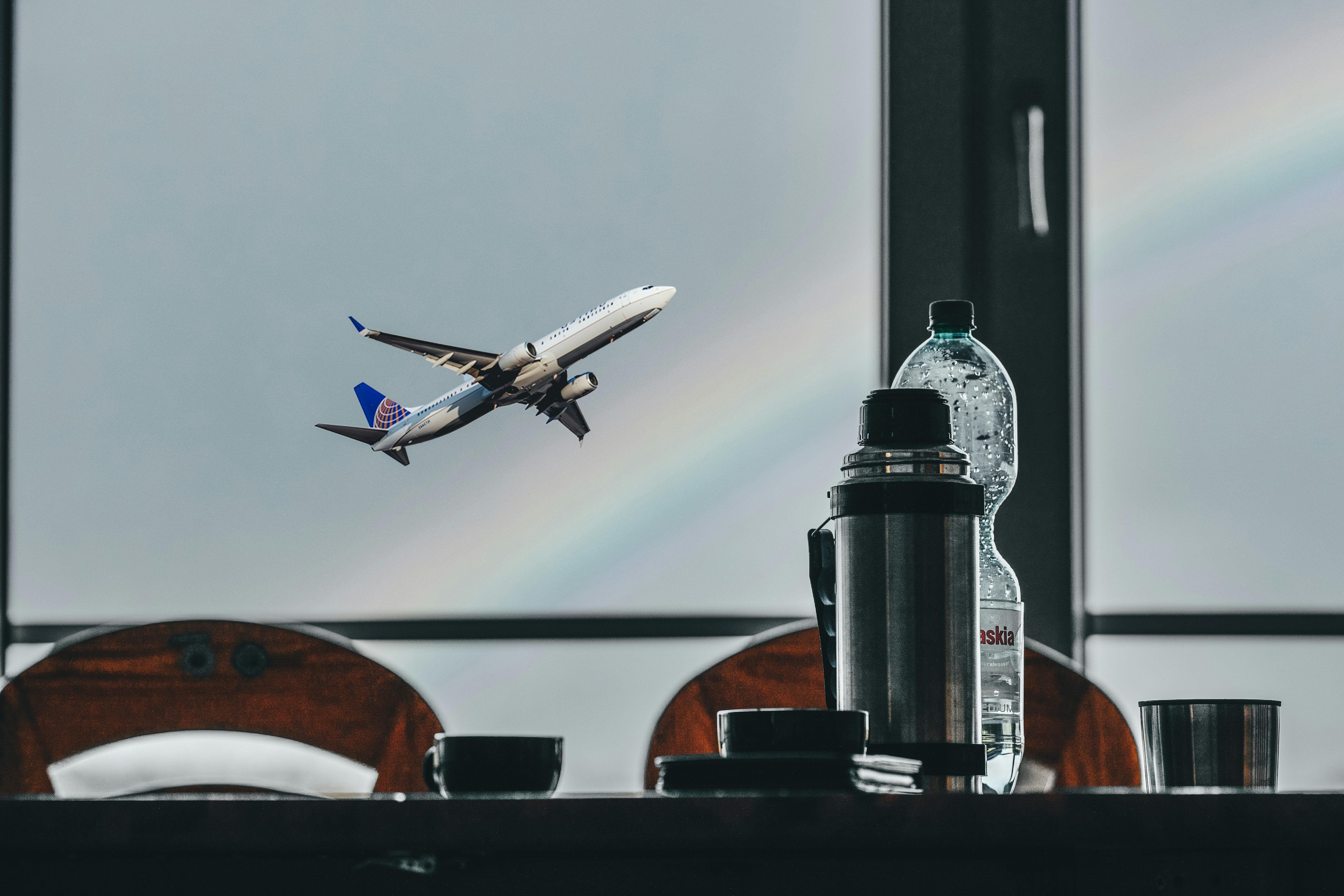 Airline in ascent visible through window behind a cluttered table with a water bottle and thermos; rainbow arc across the sky adds color to a subdued interior scene.