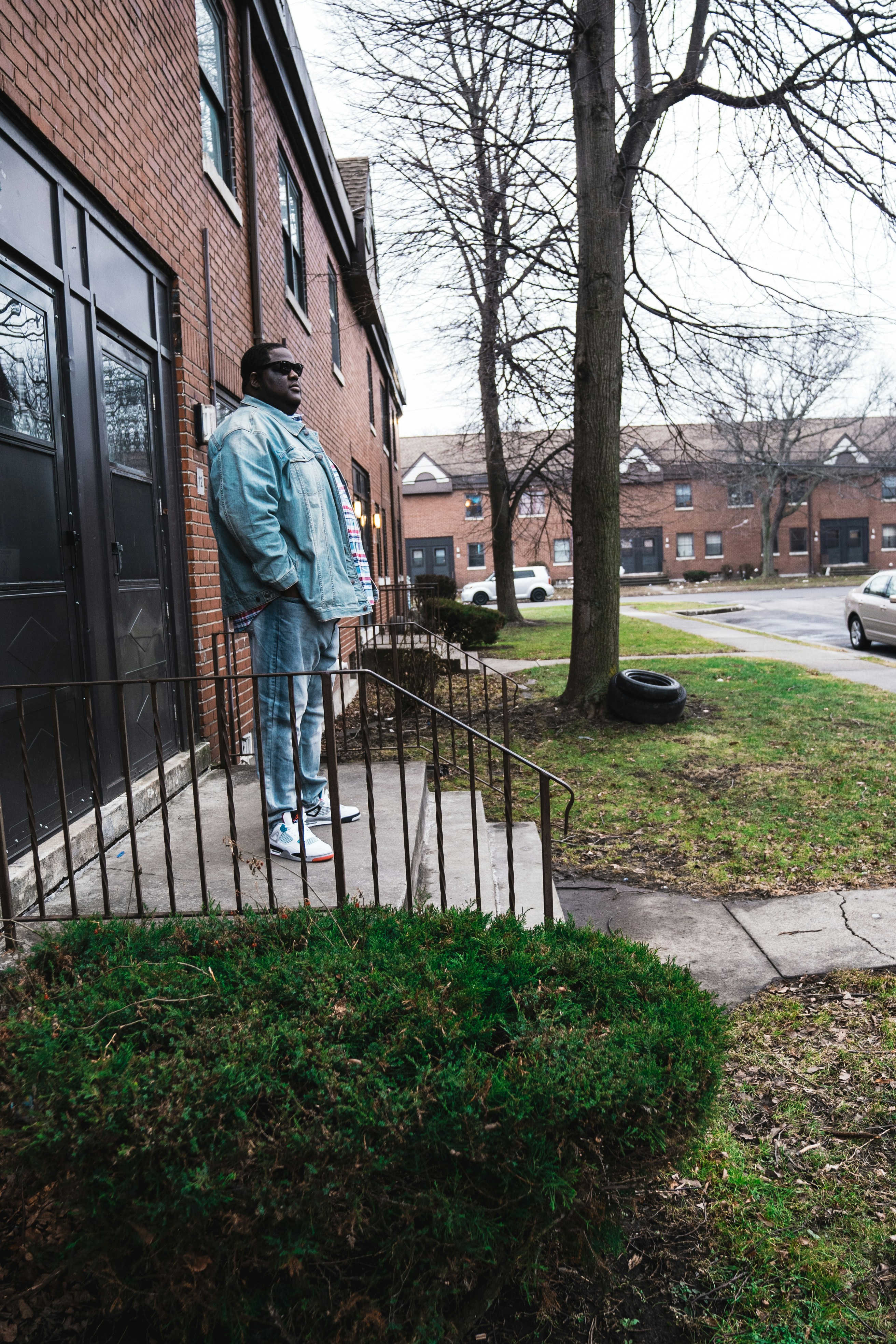 Man in denim jacket stands on a sidewalk ledge beside a brick building