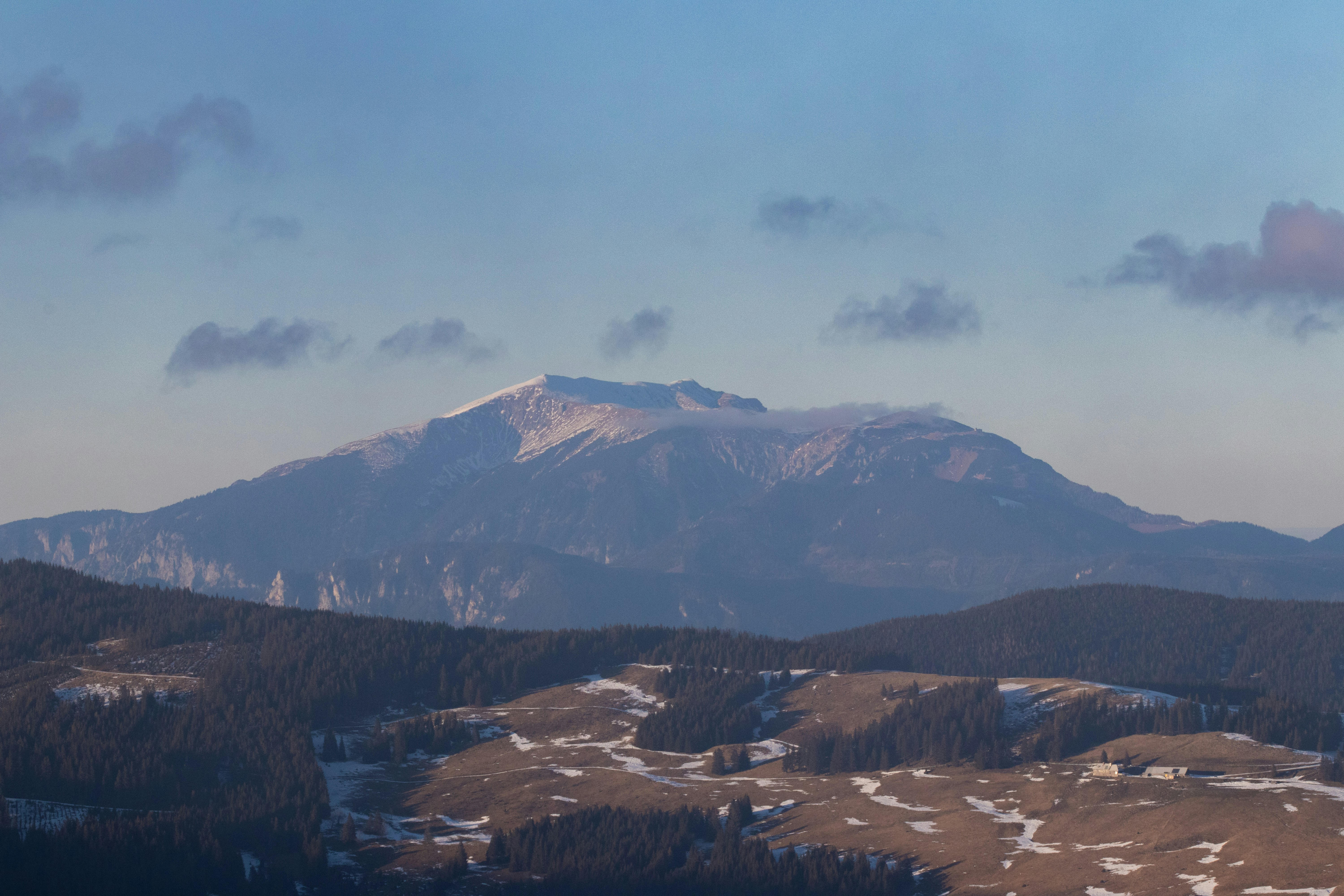 Snow-capped mountain range bathed in soft morning light, surrounded by rolling hills and patches of snow.