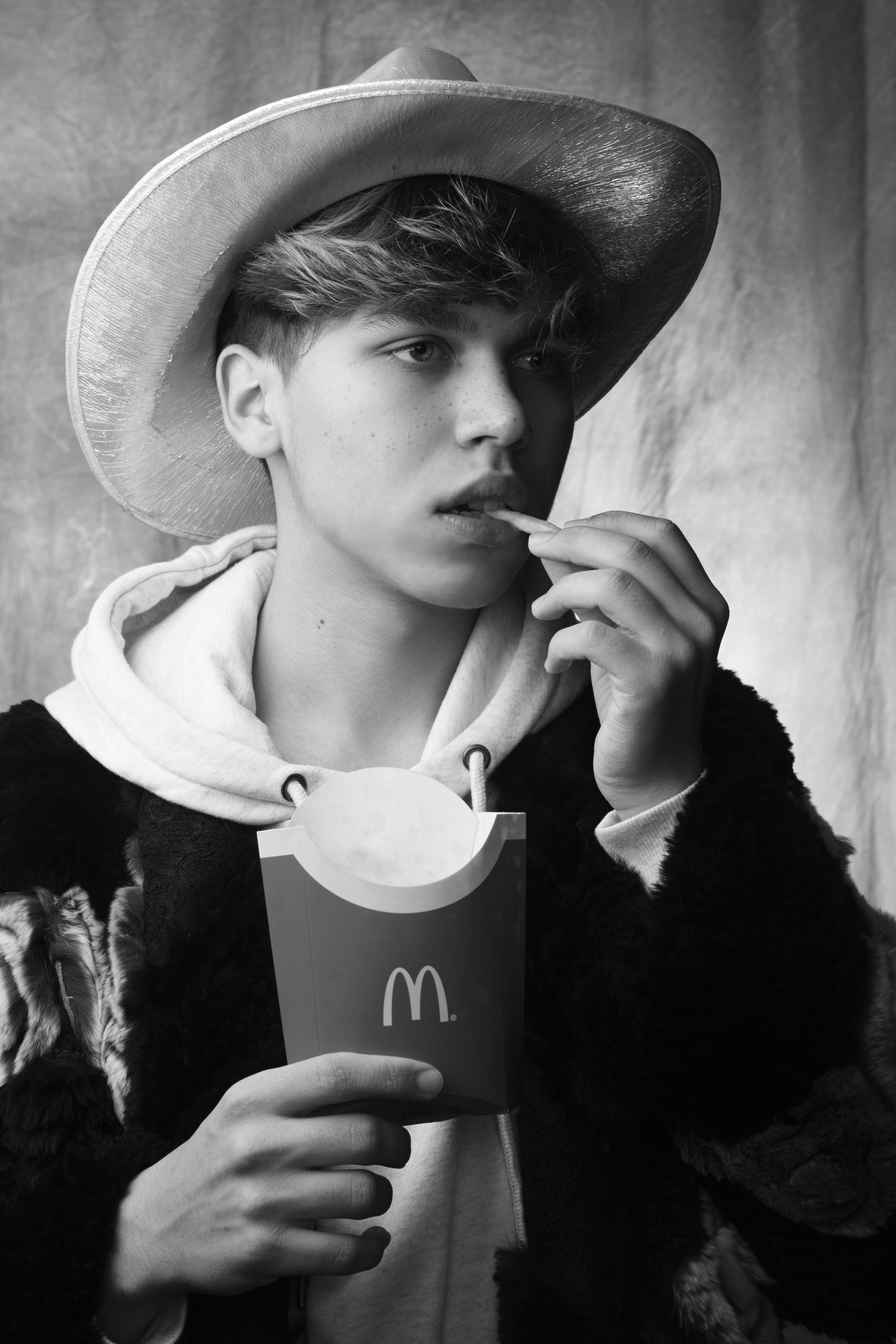 Black-and-white portrait photograph of a young man wearing a wide-brim hat and hoodie, holding a McDonald's cup while touching his lips.