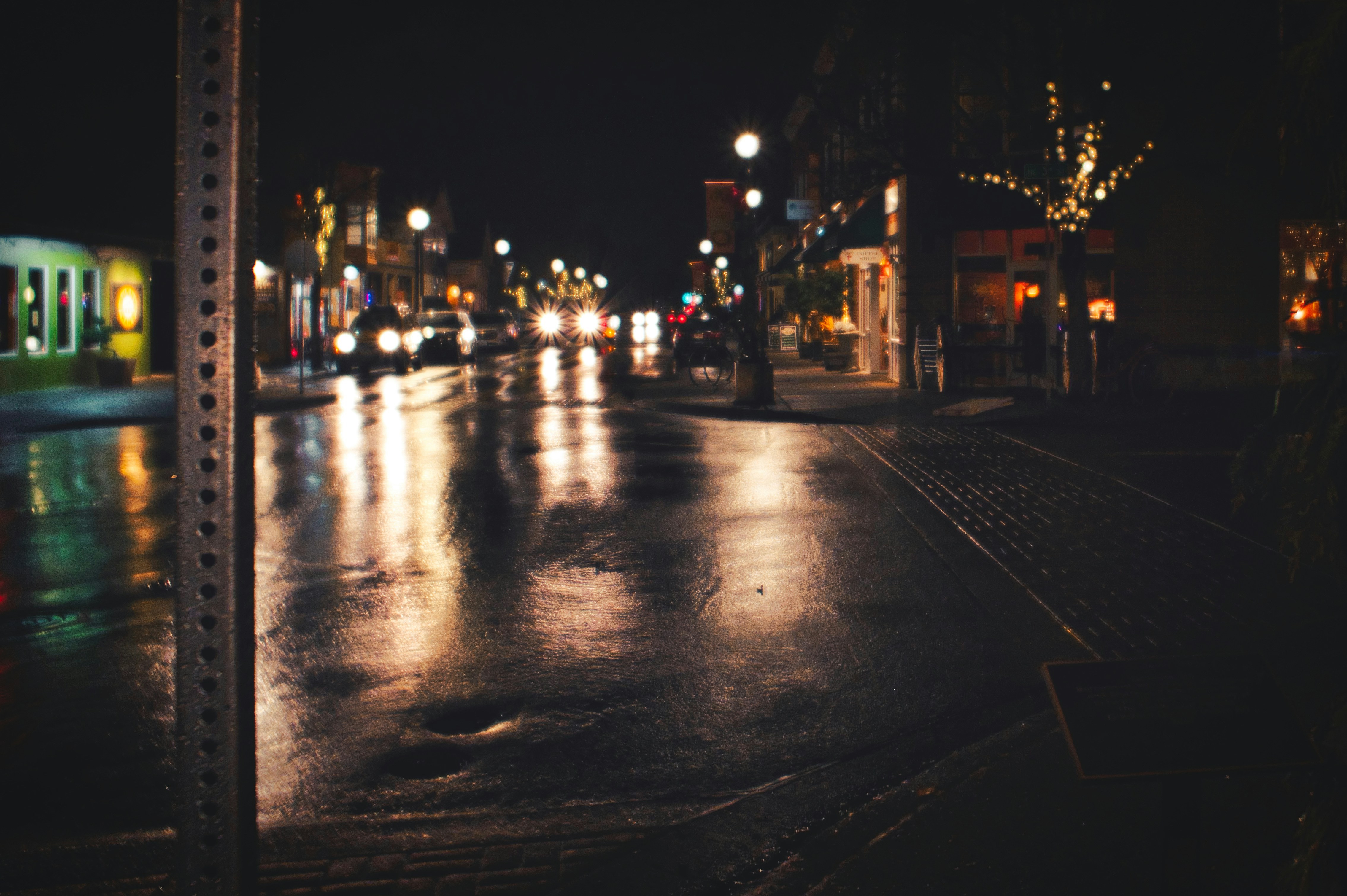 A wet street glistens under streetlights, capturing the essence of a lively urban night with cars passing by and storefronts illuminated.