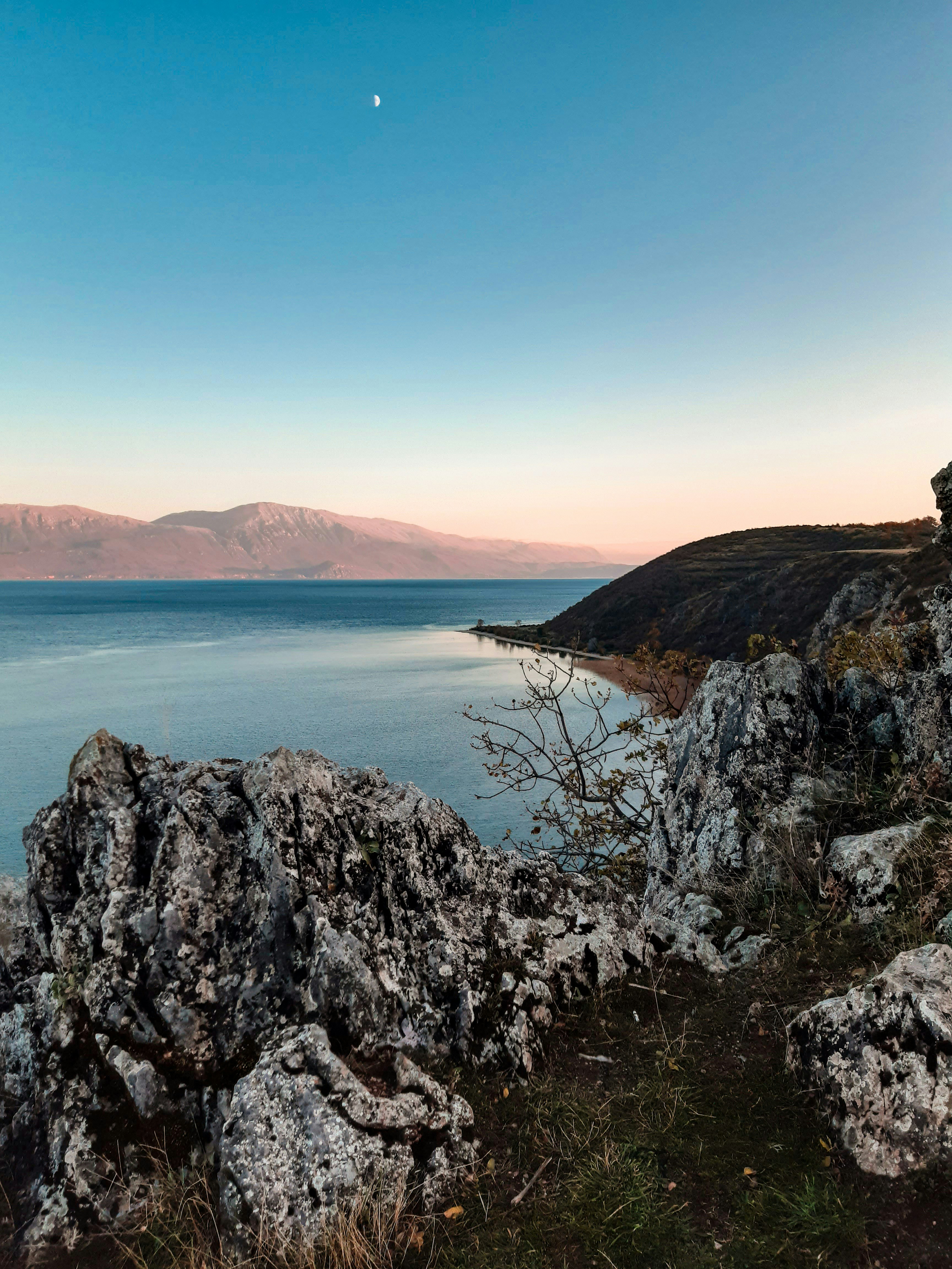 Rugged coastal rocks frame a calm sea and distant cliffs beneath a gradient sunset sky, with a crescent moon near the horizon, in a photograph.