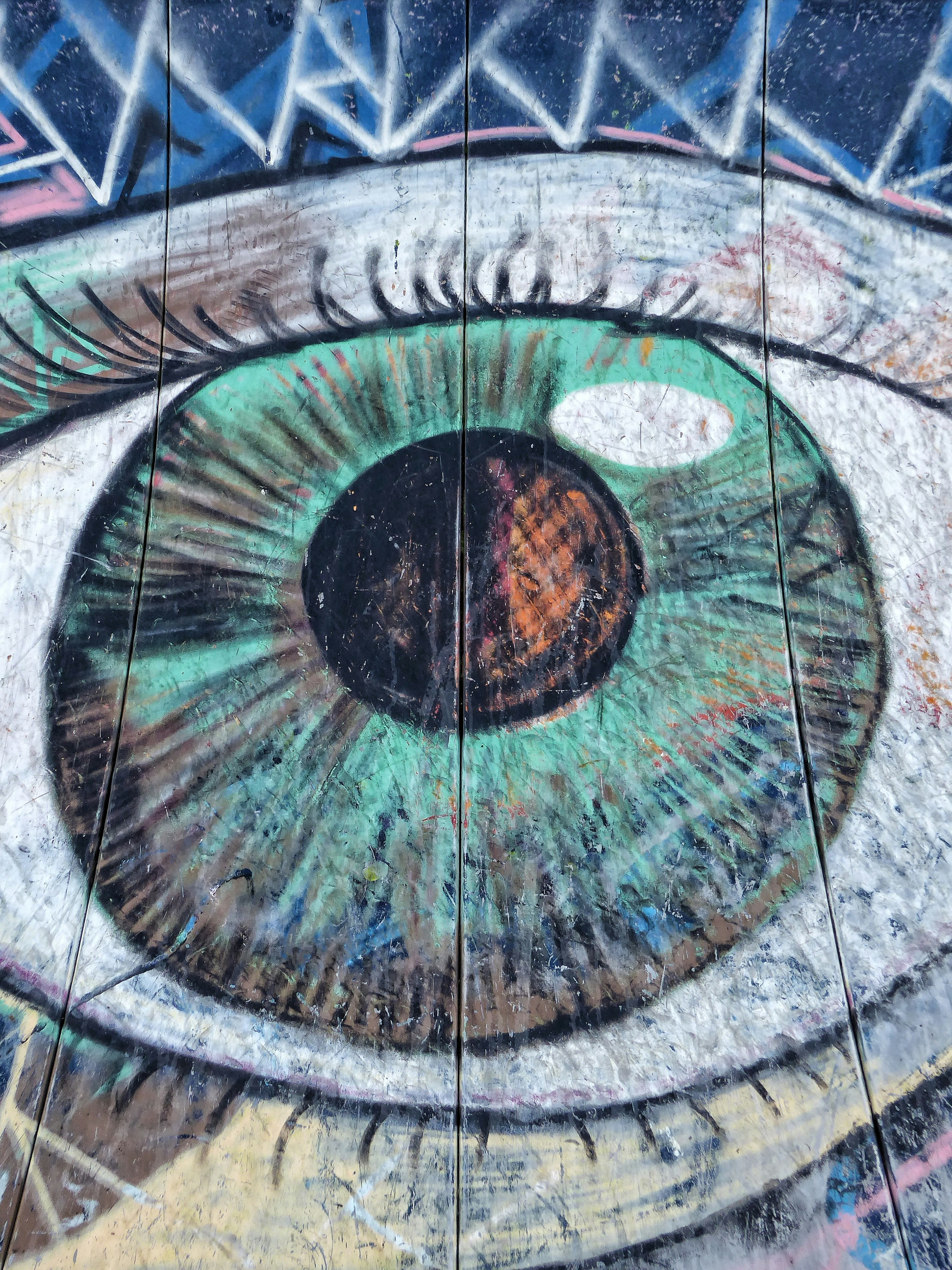 Close-up of a vivid graffiti eye with a bright green iris and textured paint on a wooden surface. A bold street-art detail emphasizing color and grit.