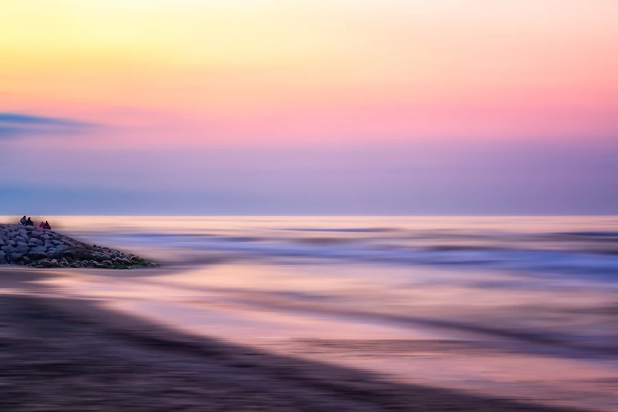 Long-exposure seascape with a rocky breakwater on the left, blurred waves, and a pastel sunset sky fading from gold to pink and lavender.