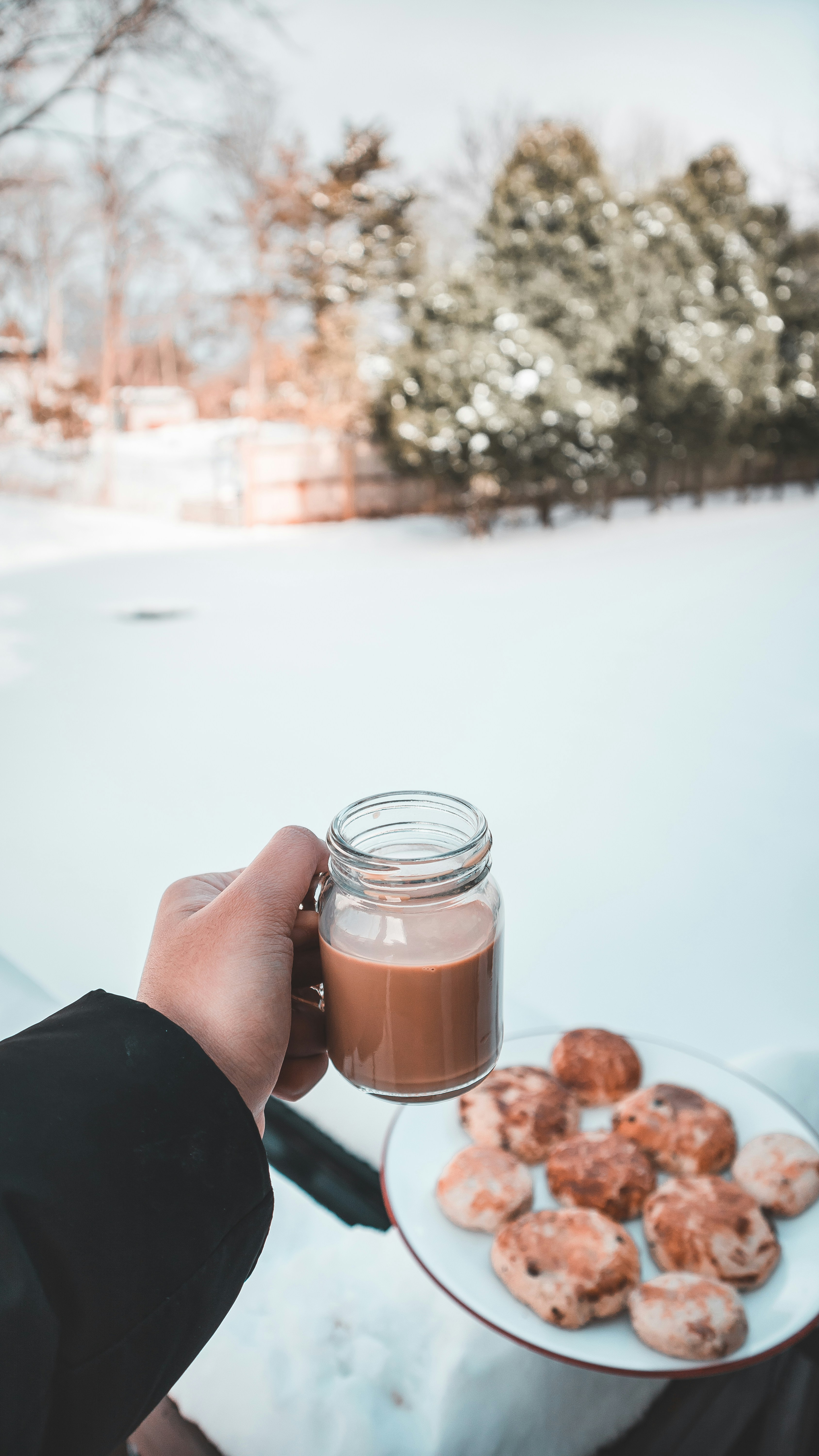 Hand holding a jar of chai with a plate of cookies against a snowy backyard backdrop.
