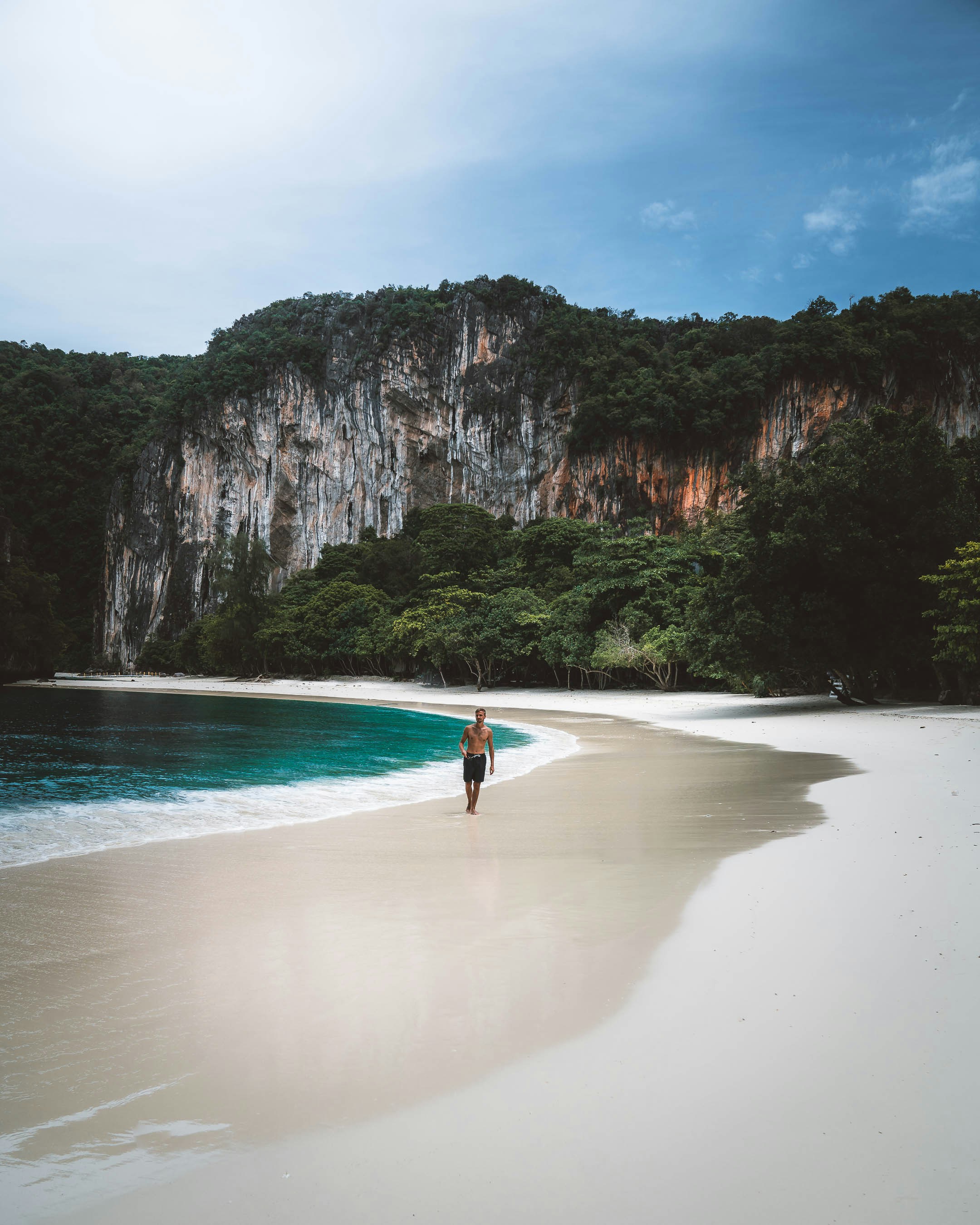 Person walking along a deserted white sand beach with towering cliffs and lush greenery under a partly cloudy sky.