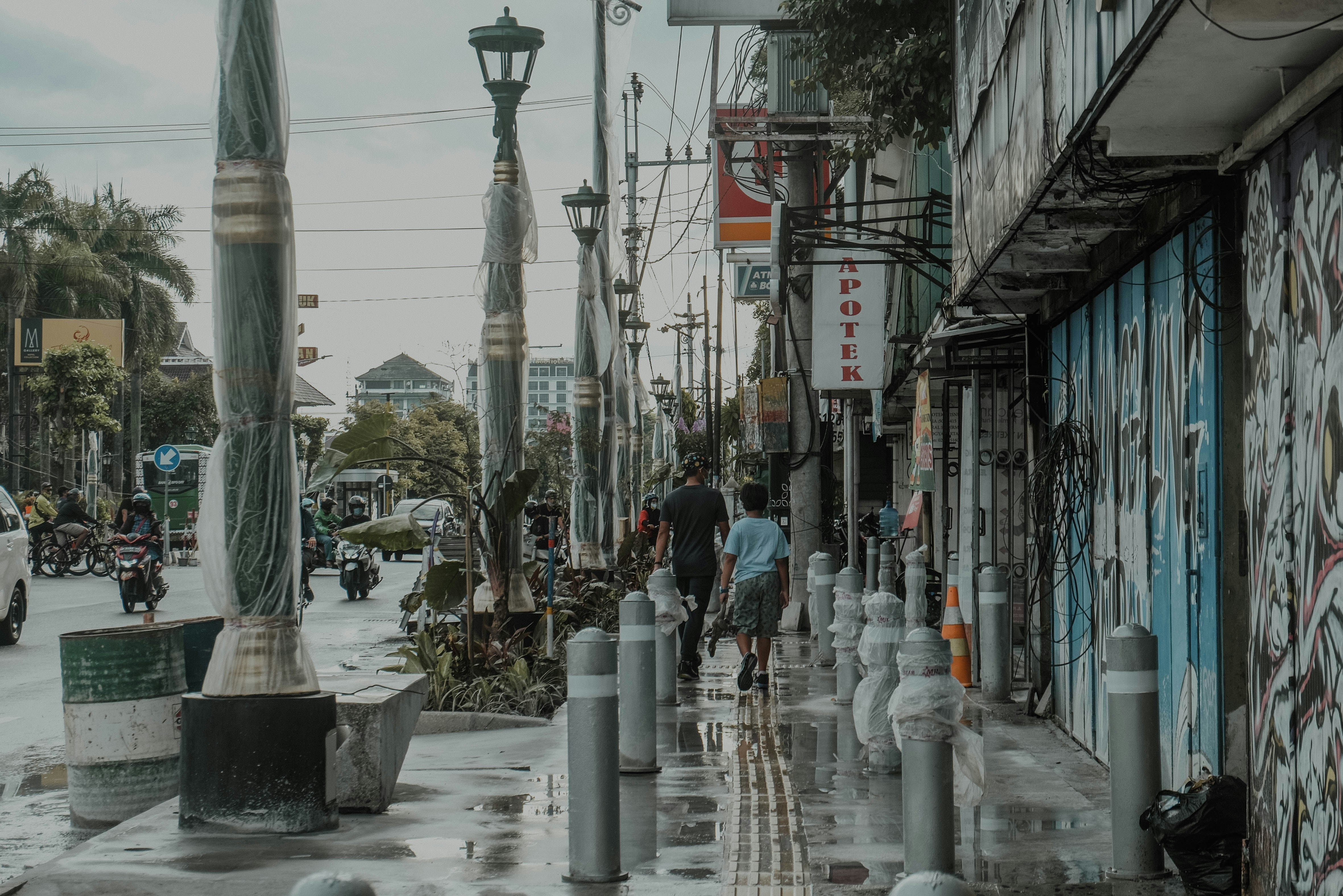 Pedestrians navigate a graffiti-lined sidewalk in an urban setting under overcast skies.
