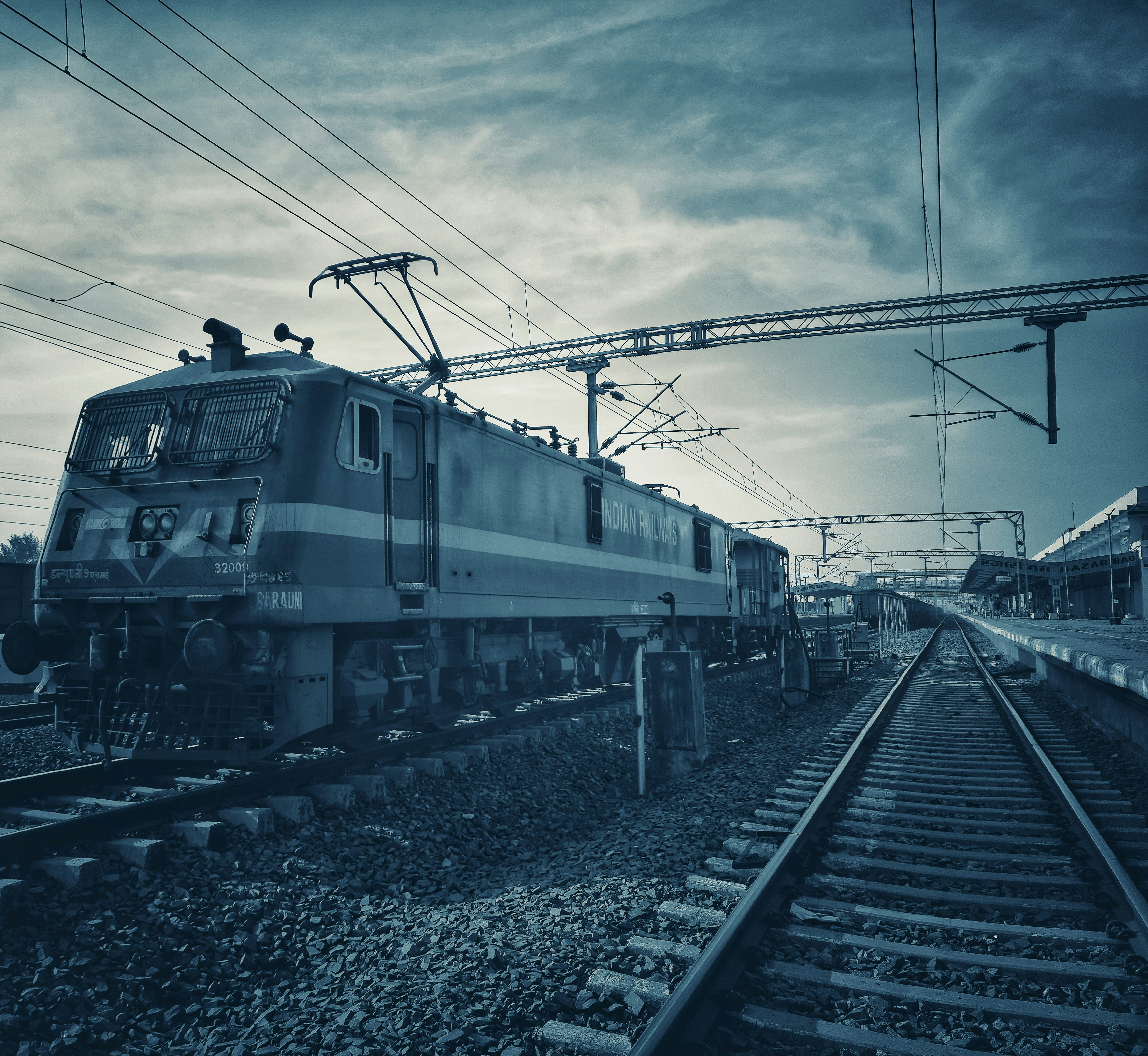 Blue-toned photograph of a diesel freight locomotive in a quiet rail yard, with overhead electric lines and parallel tracks. The composition emphasizes the engine's mass against converging rails.