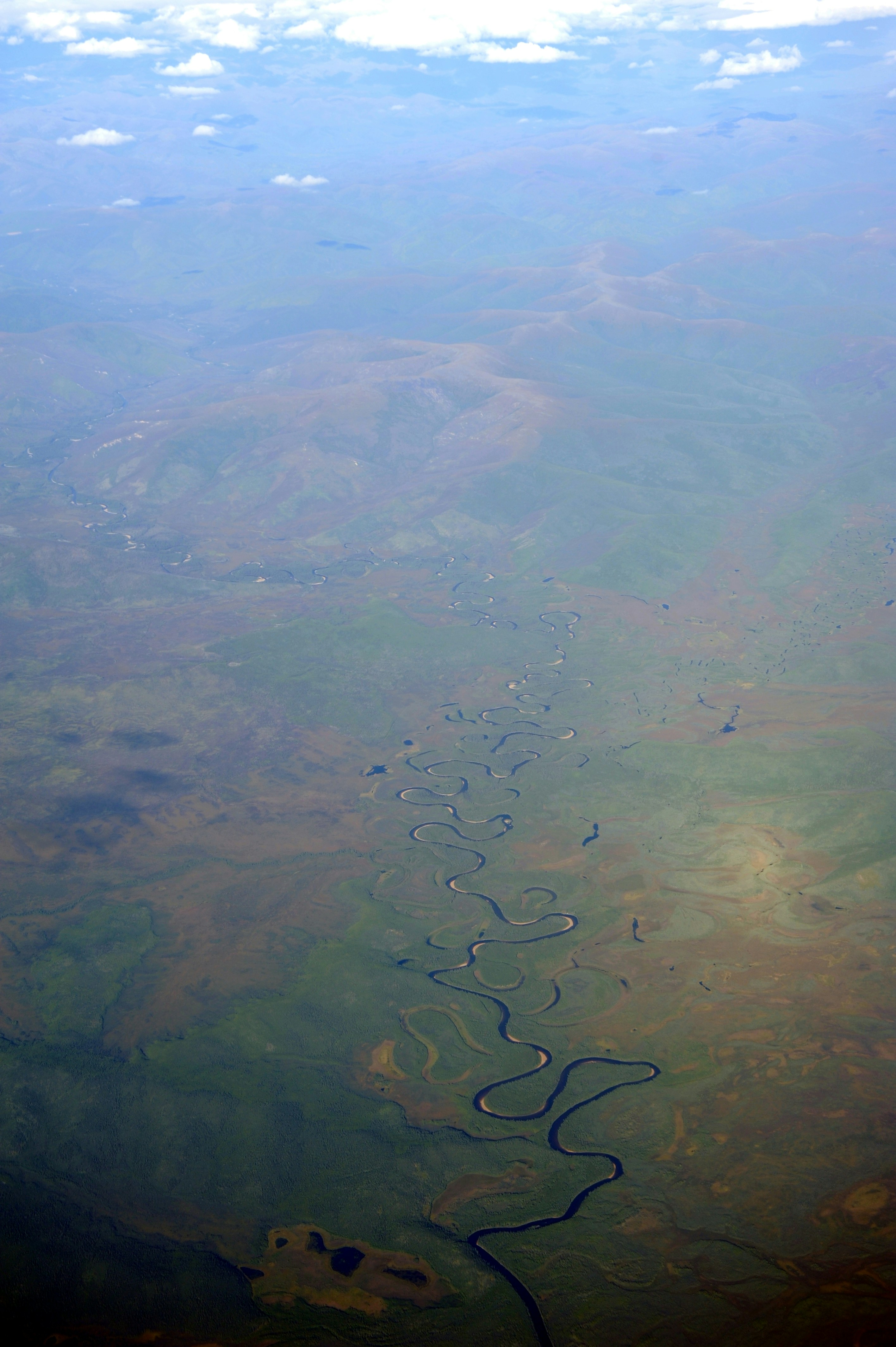 Aerial view of a meandering river cutting through a vast green landscape, showcasing the intricate patterns of nature. Clouds scatter across the blue sky above.