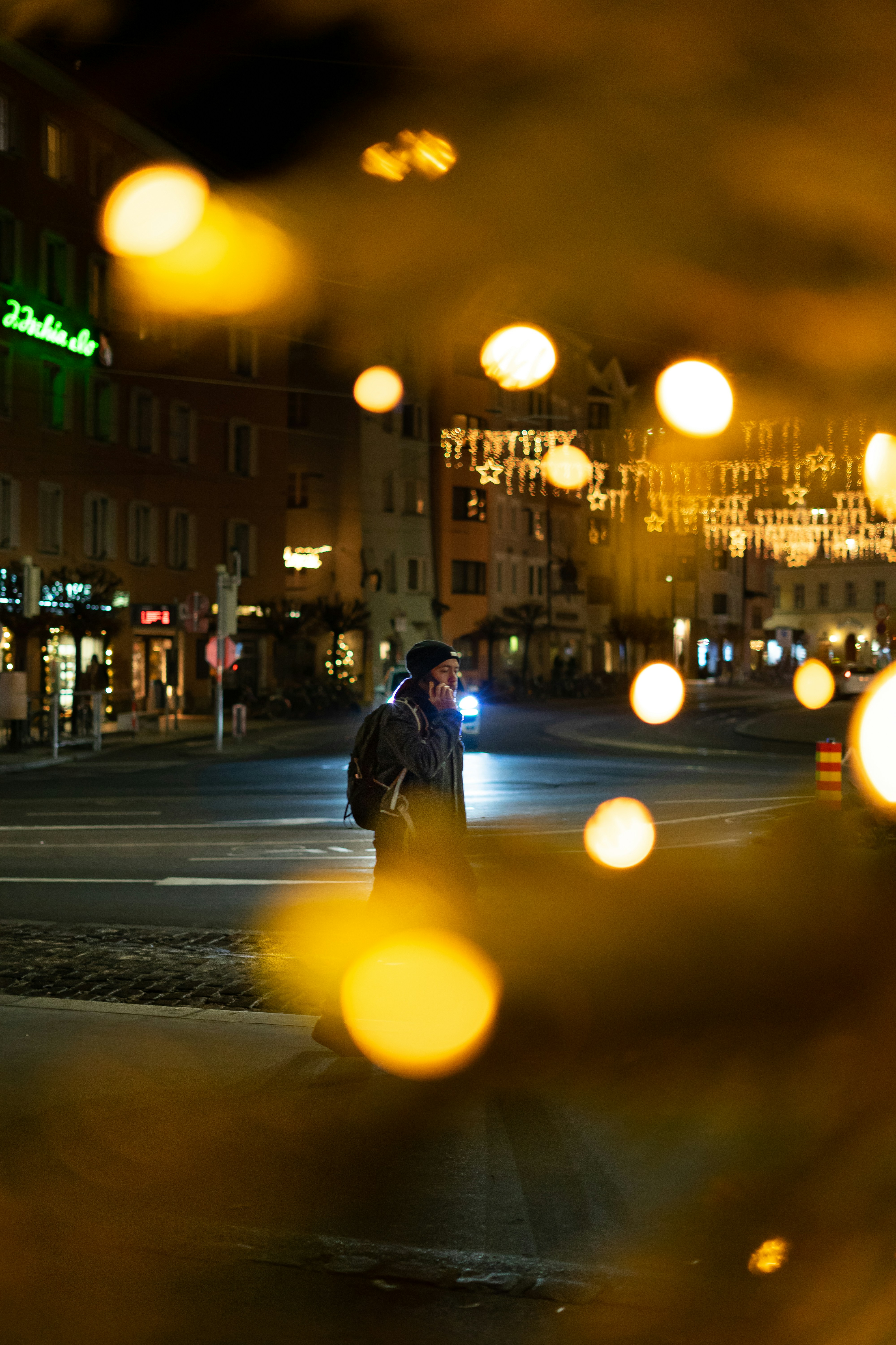 A lone figure stands on a city street, illuminated by festive lights, while soft bokeh circles frame the scene. The atmosphere conveys a sense of quiet reflection amidst holiday cheer.