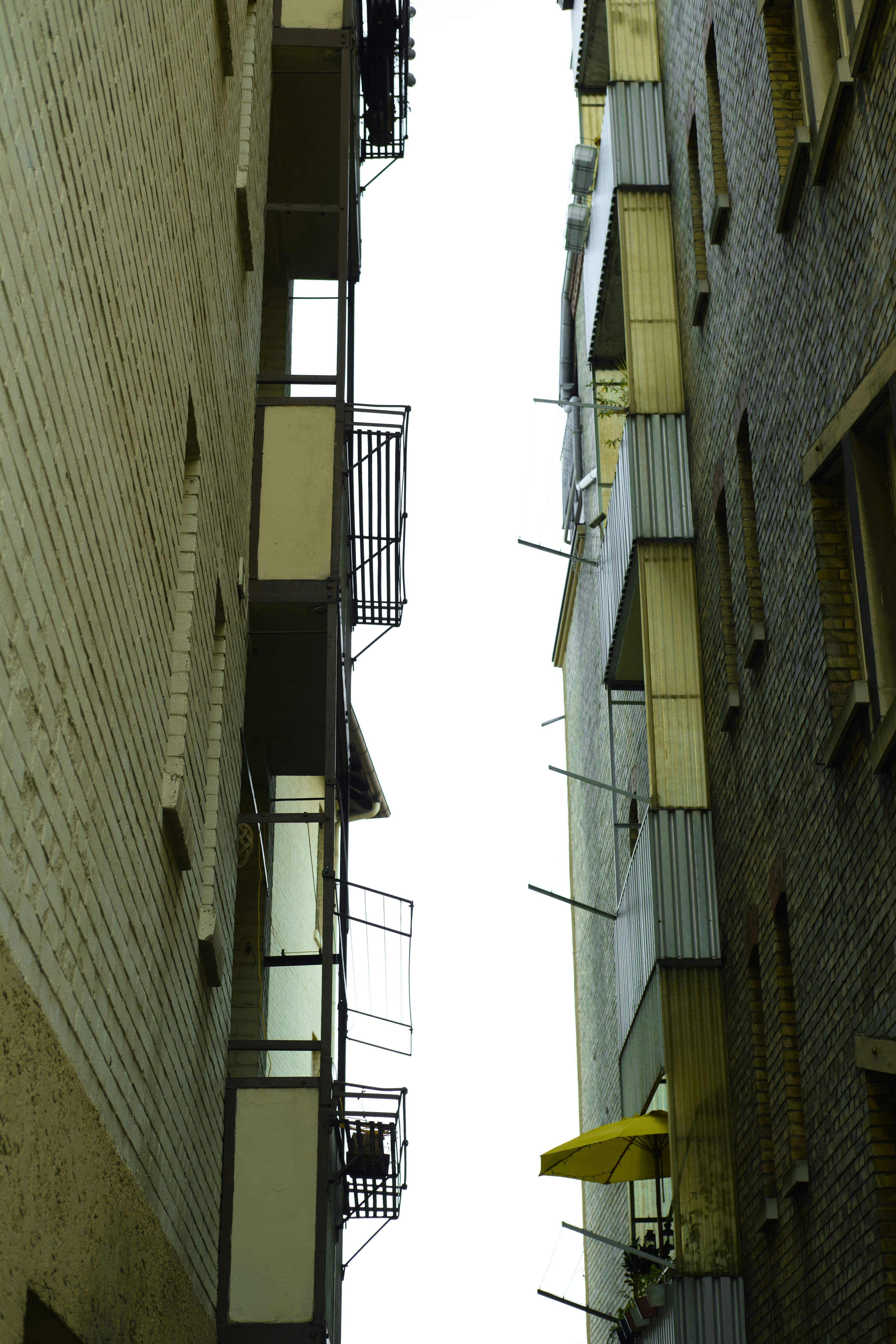 Narrow alleyway flanked by towering buildings, featuring balconies and a yellow umbrella peeking through the space above.