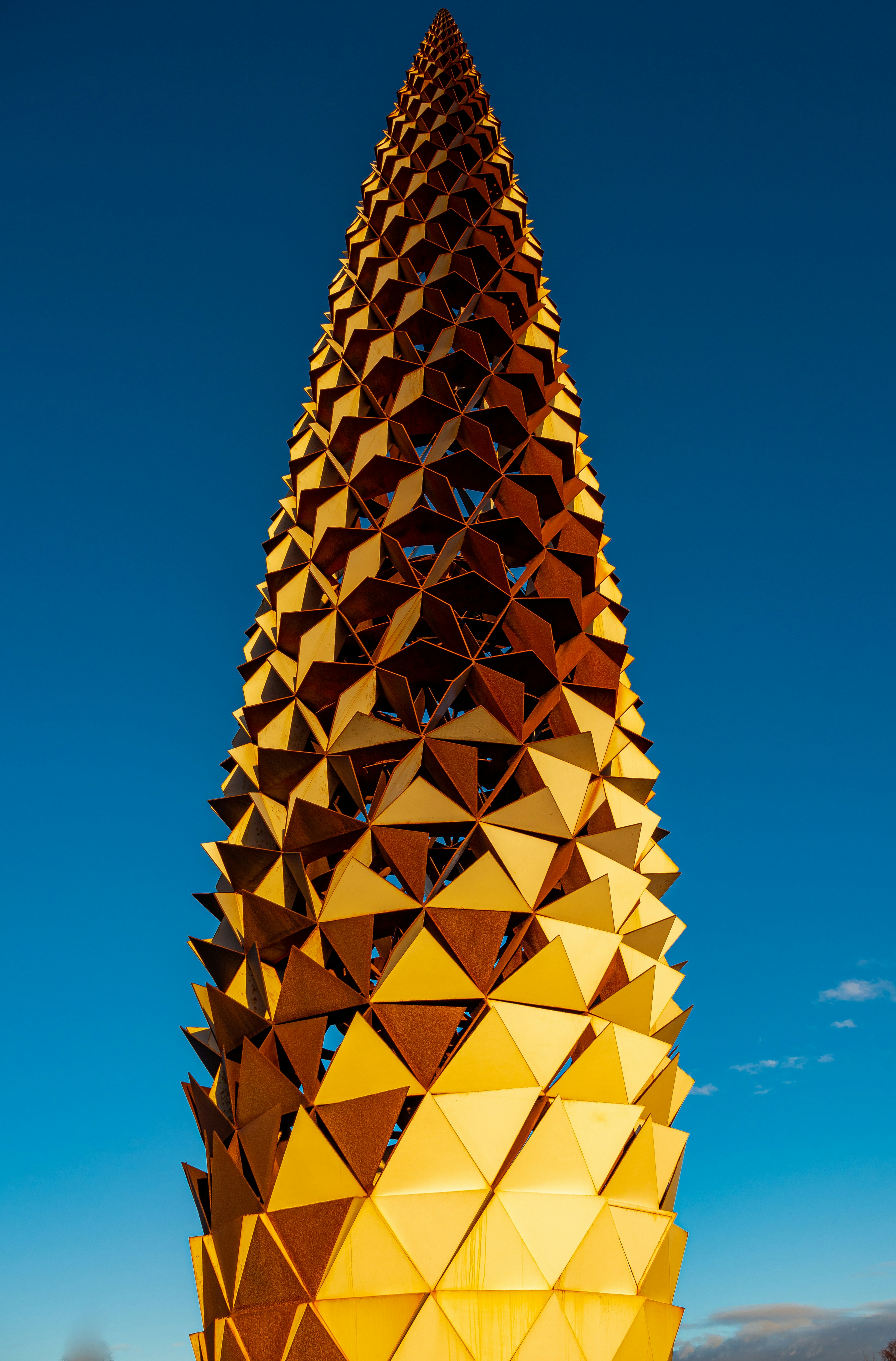A striking golden sculpture with intricate geometric patterns reaching towards the sky against a clear blue backdrop.