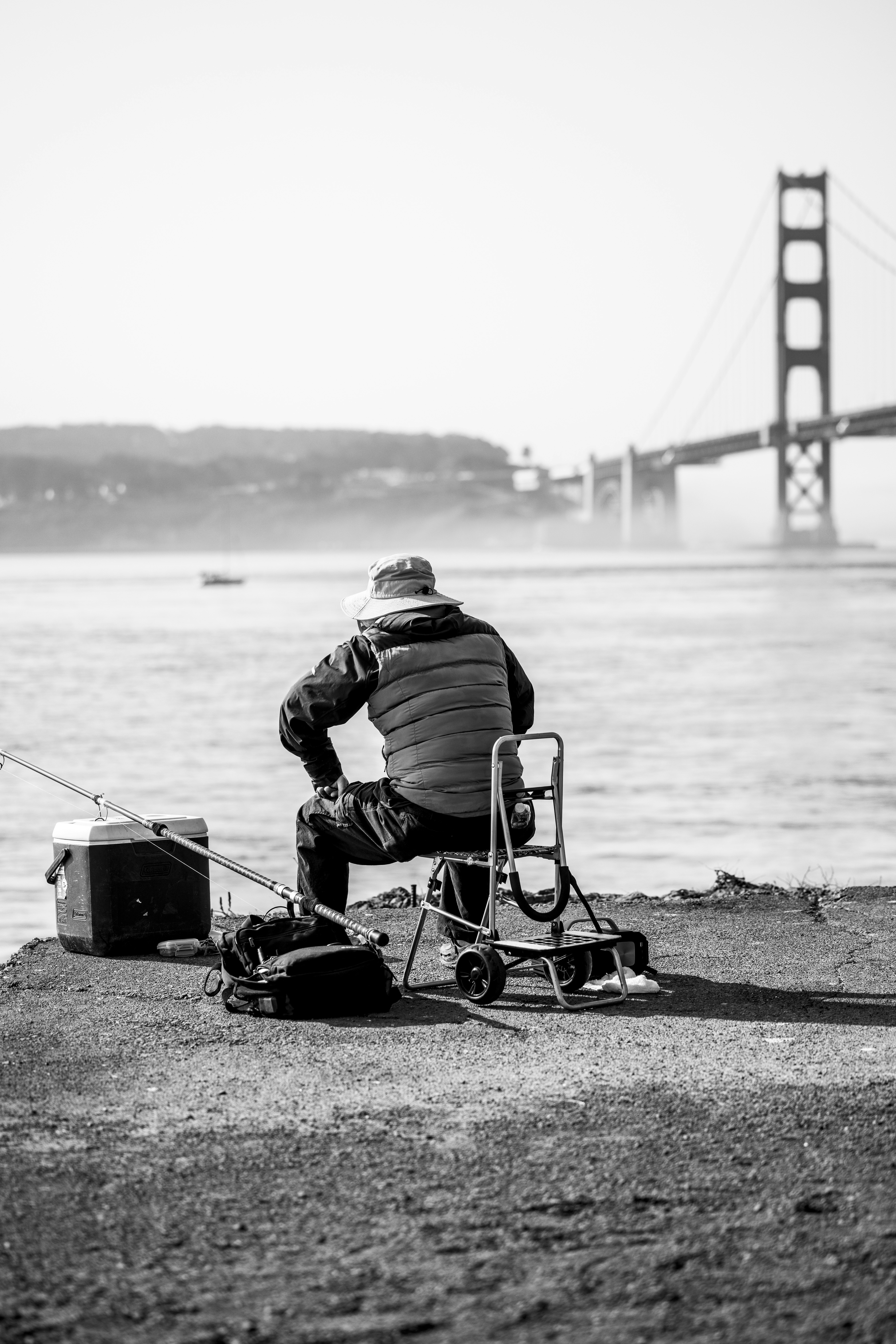 Fisherman seated on a dock, casting a line into the water with the Golden Gate Bridge in the background. Black and white tones enhance the serene atmosphere.