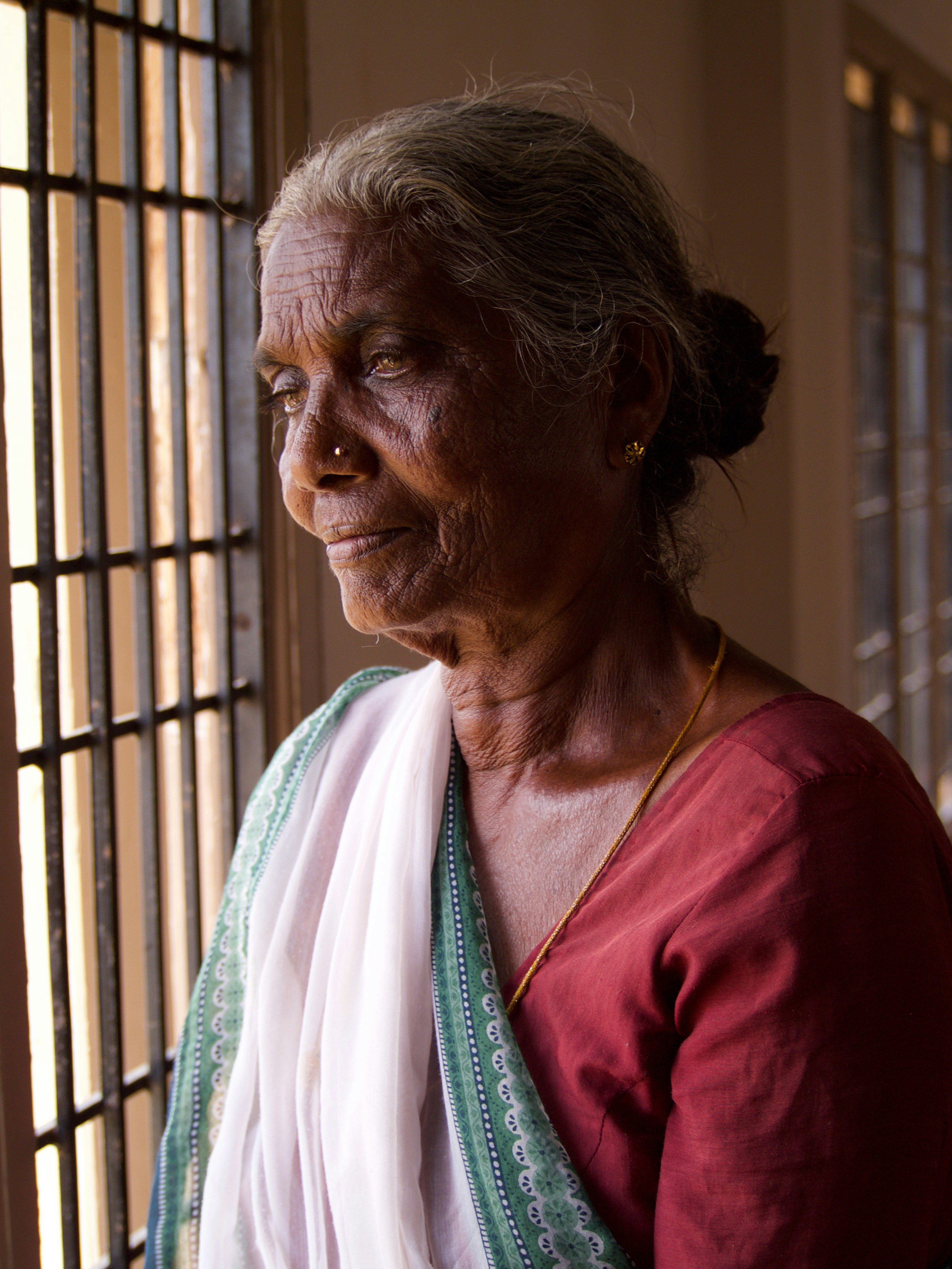 Elderly woman gazing thoughtfully out of a window, bathed in soft light, showcasing her serene expression and traditional attire.