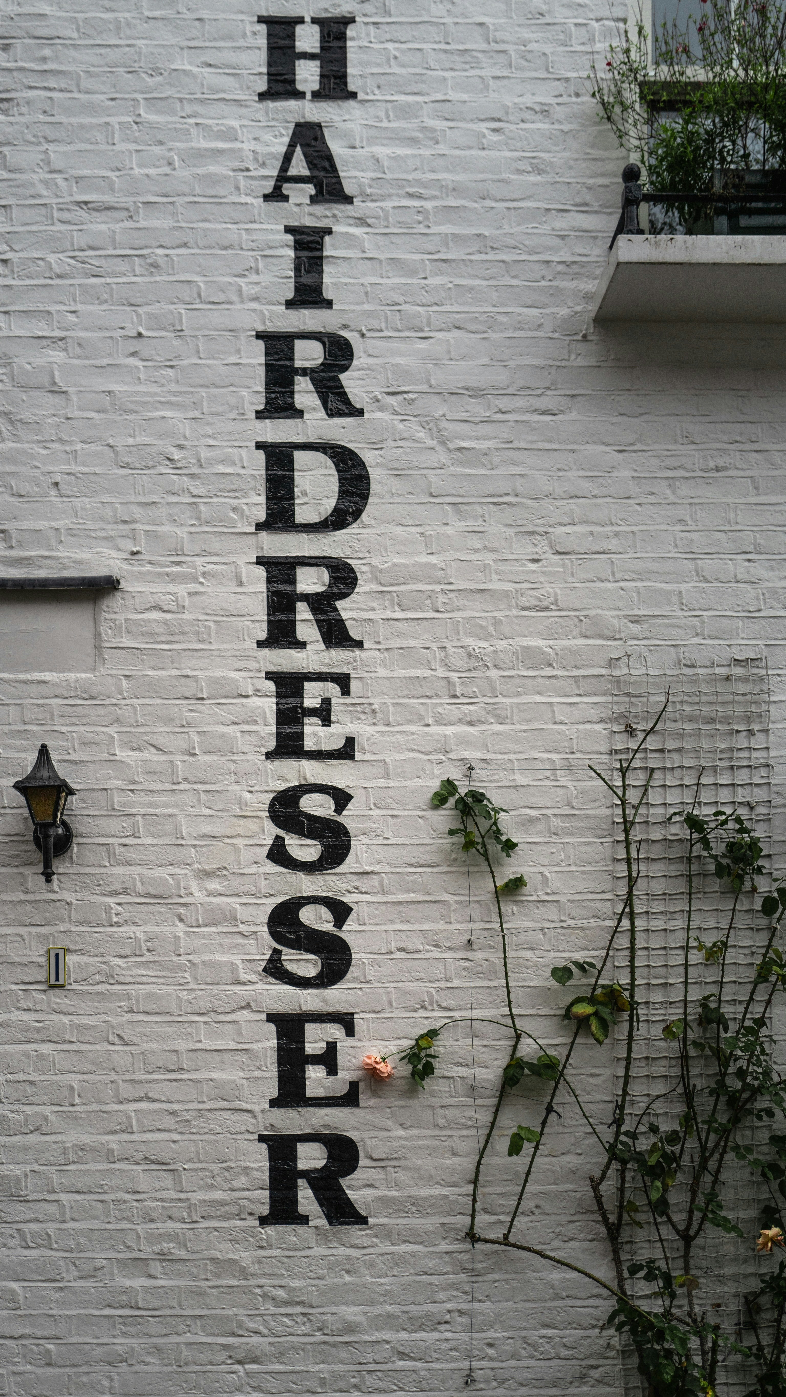 A bold 'HAIRDRESSER' sign painted vertically on a textured white wall, accompanied by delicate greenery and a hint of floral decor.
