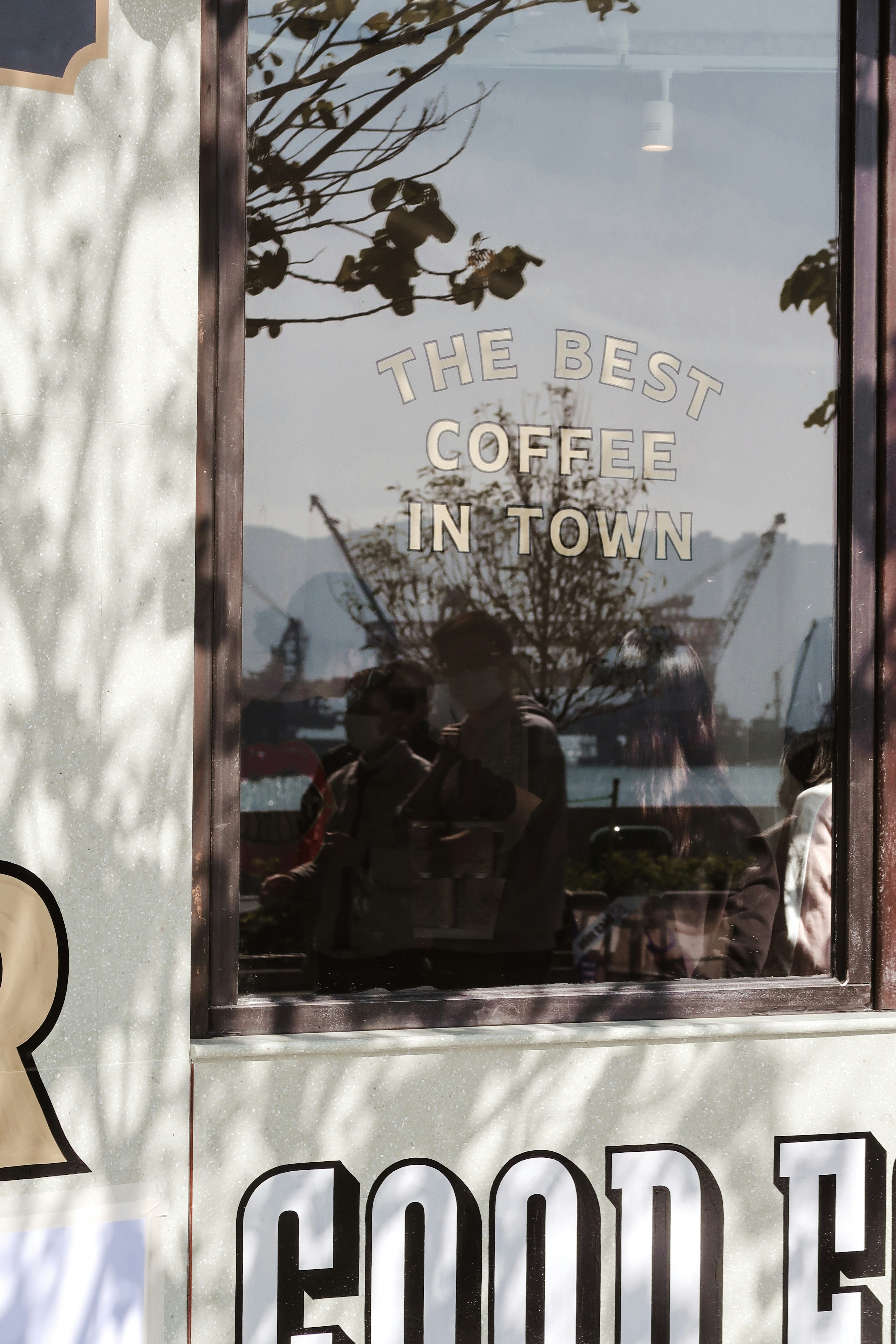 The window of a coffee shop showcasing the words 'THE BEST COFFEE IN TOWN' with reflections of passersby and a scenic backdrop.