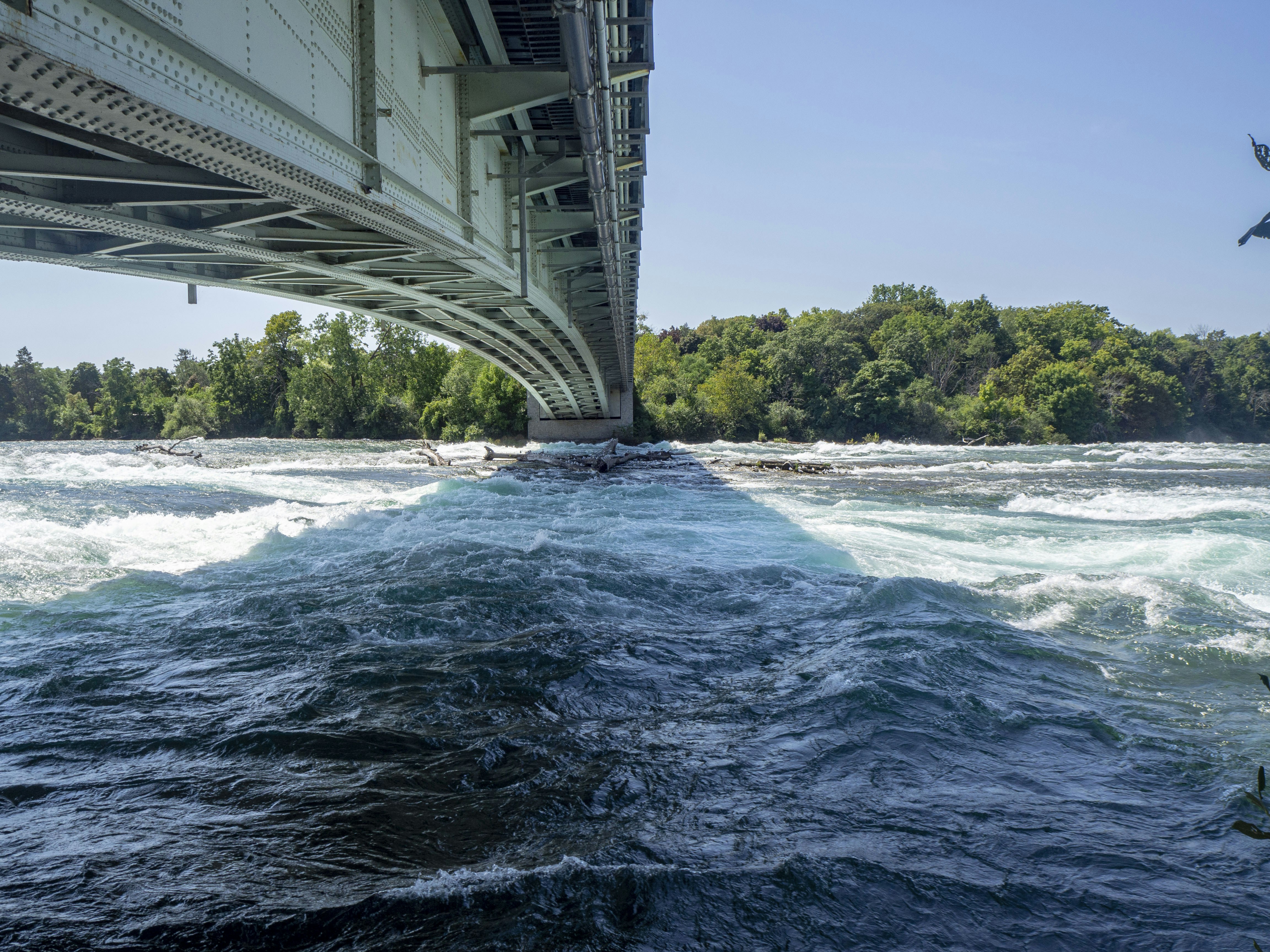 Swift river currents swirl beneath a robust bridge, framed by lush greenery on the banks. The interplay of light and shadow highlights the dynamic movement of the water.