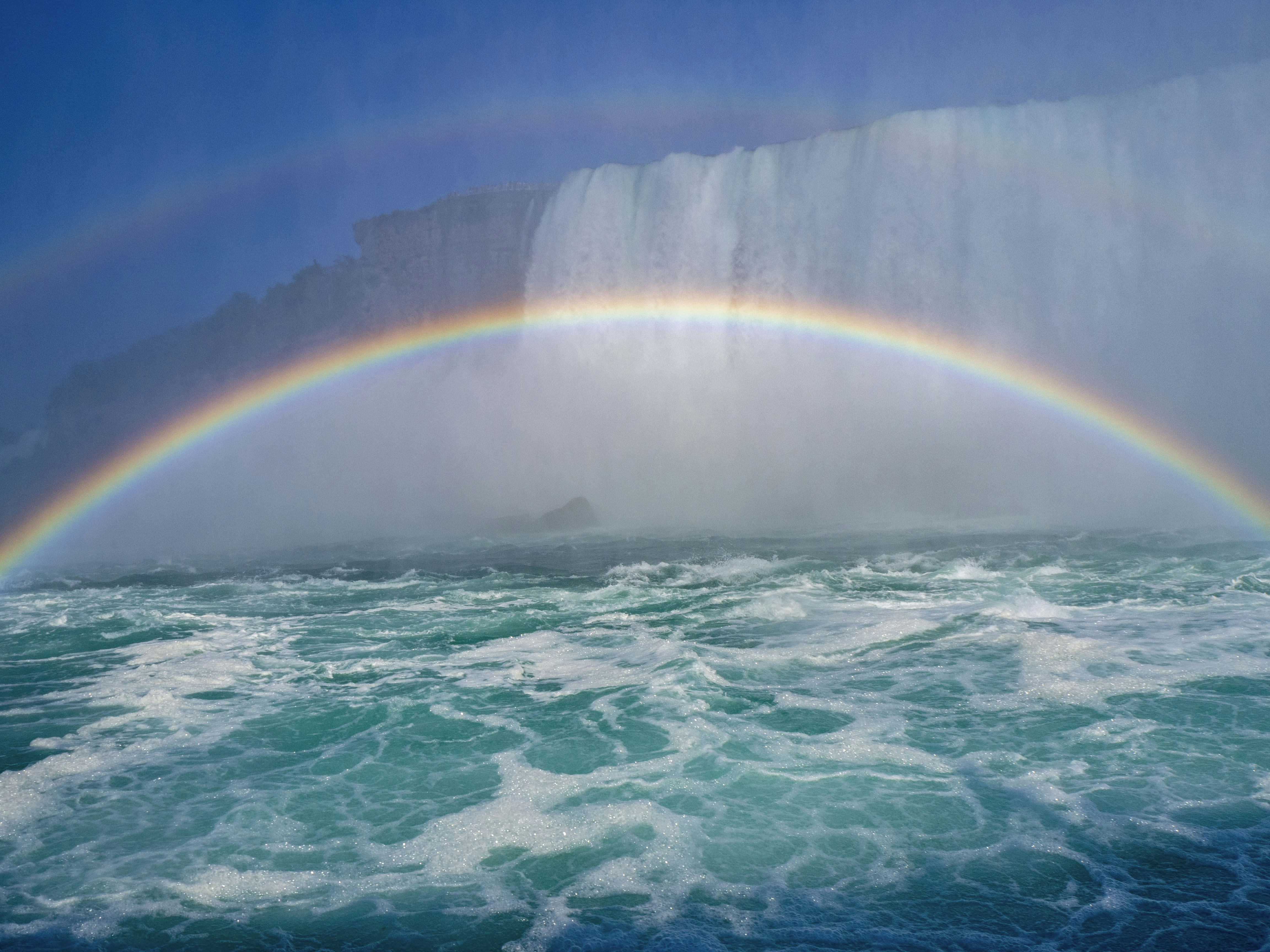Double rainbow arches above the misty cascade of Niagara Falls against a vibrant blue sky.