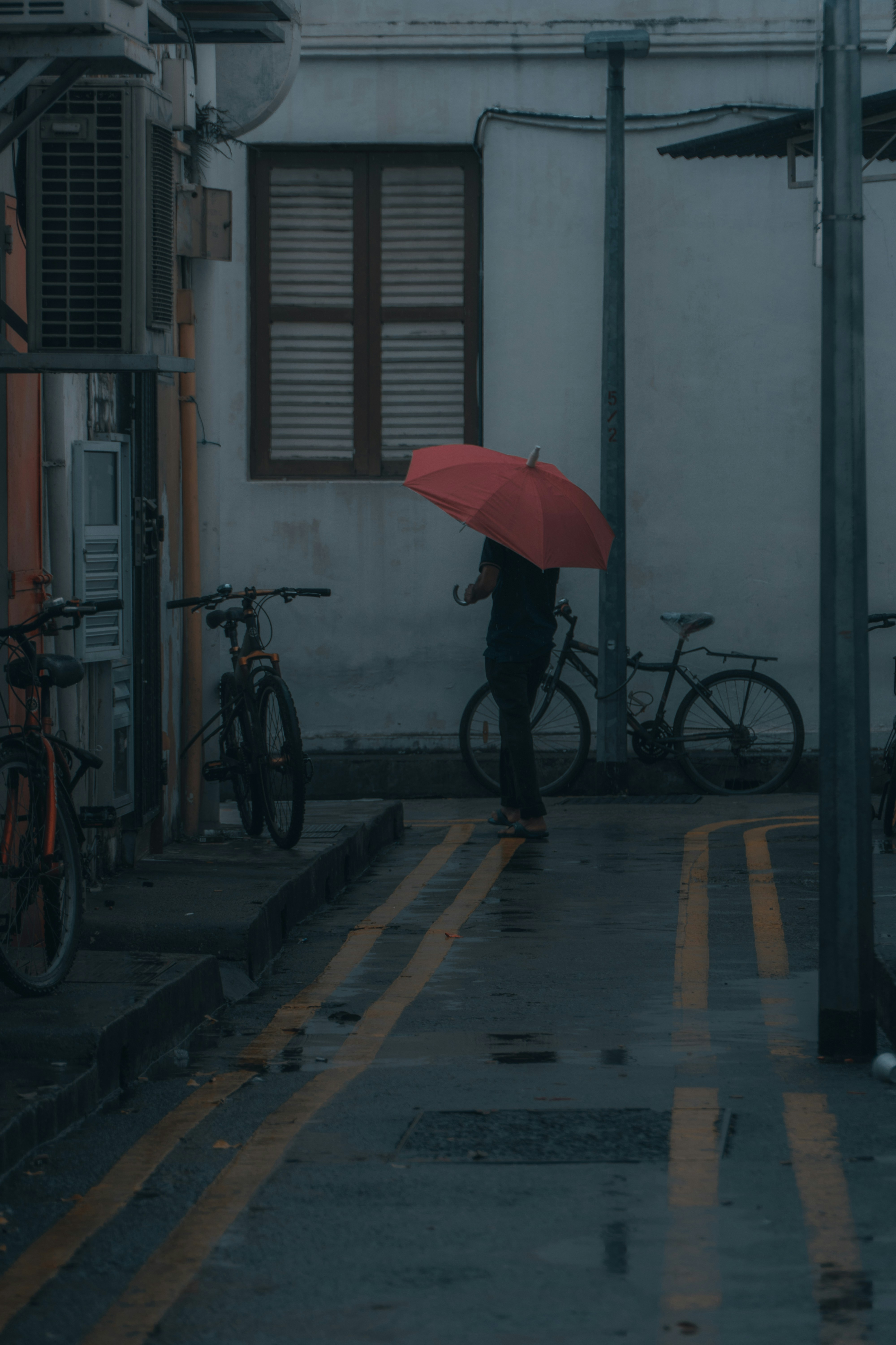 A solitary figure with a red umbrella navigates a wet alley lined with bicycles, evoking a sense of quiet reflection in a bustling urban environment.