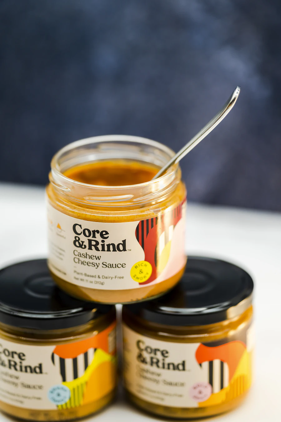 Three Core & Rind cashew cheesy sauce jars stacked on a white surface, with a spoon in the open top jar. The label is clearly visible against a dark blue background.