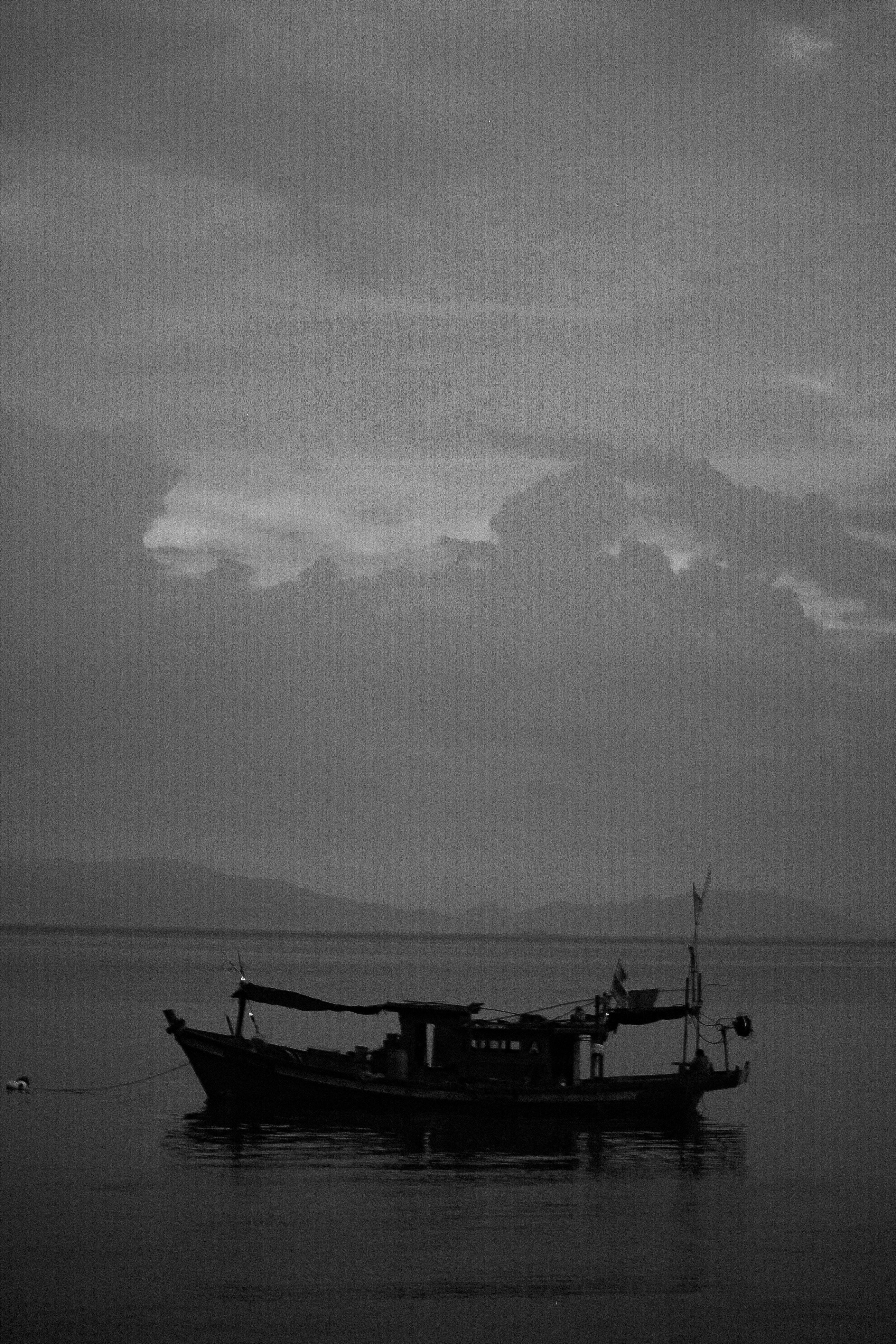 A solitary fishing boat anchored on calm waters, surrounded by a serene, cloudy sky. The monochrome tones enhance the tranquil atmosphere.