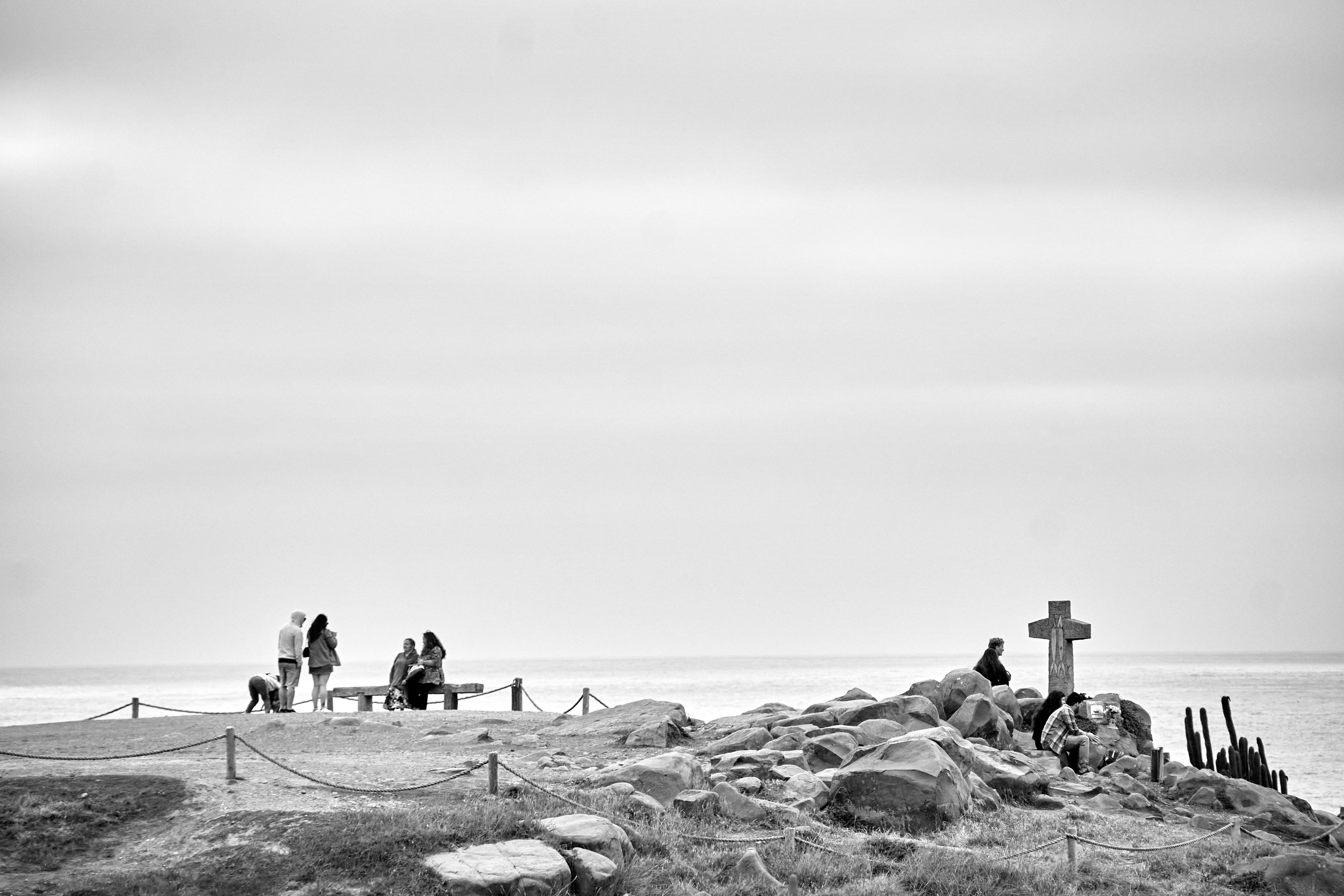 Group of people gathered near a rocky shoreline, with a cross in the foreground, capturing a serene moment by the ocean. Black and white tones enhance the emotional depth.
