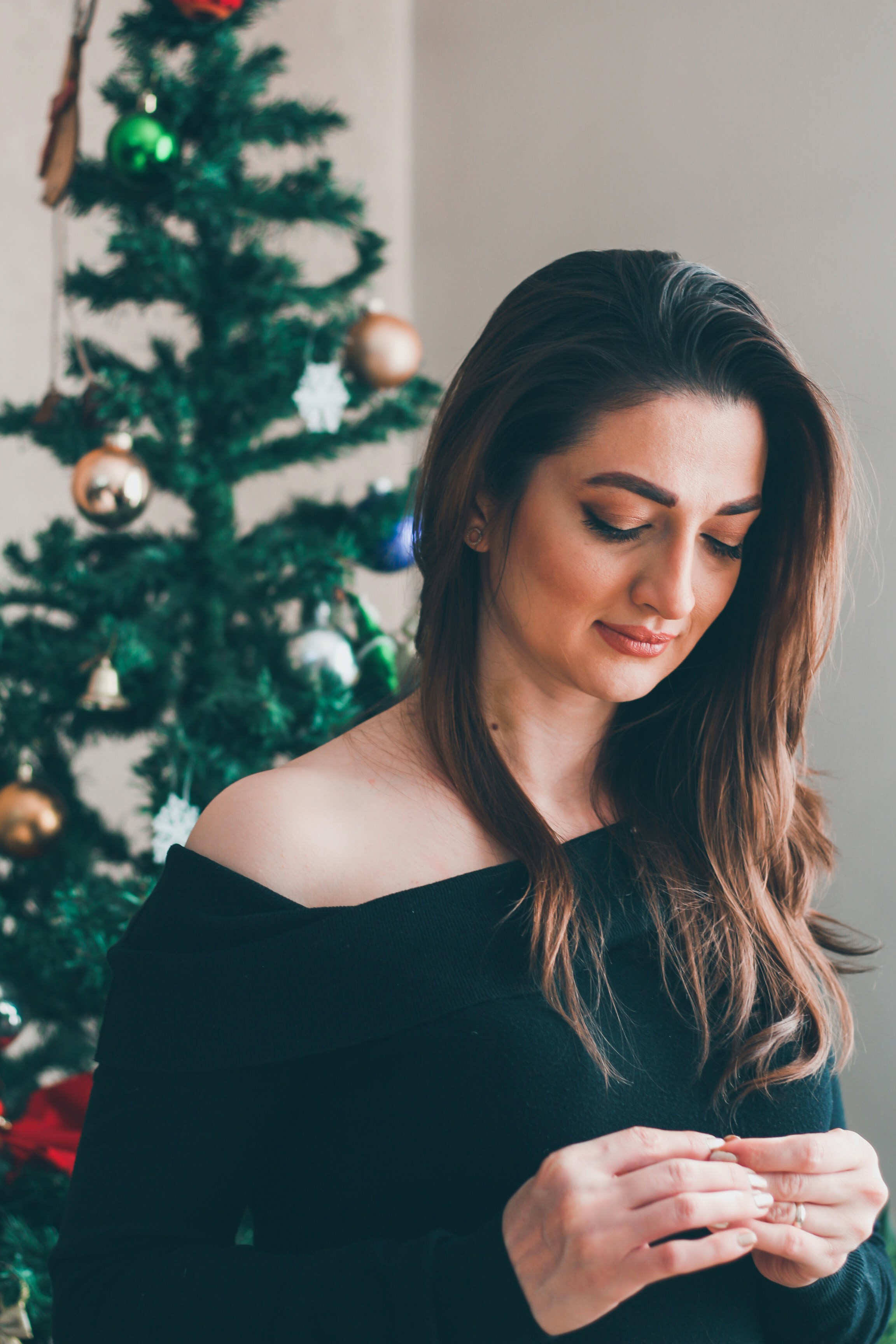 Woman in a black off-shoulder top gazes down thoughtfully, adorned by a festive Christmas tree in the background.