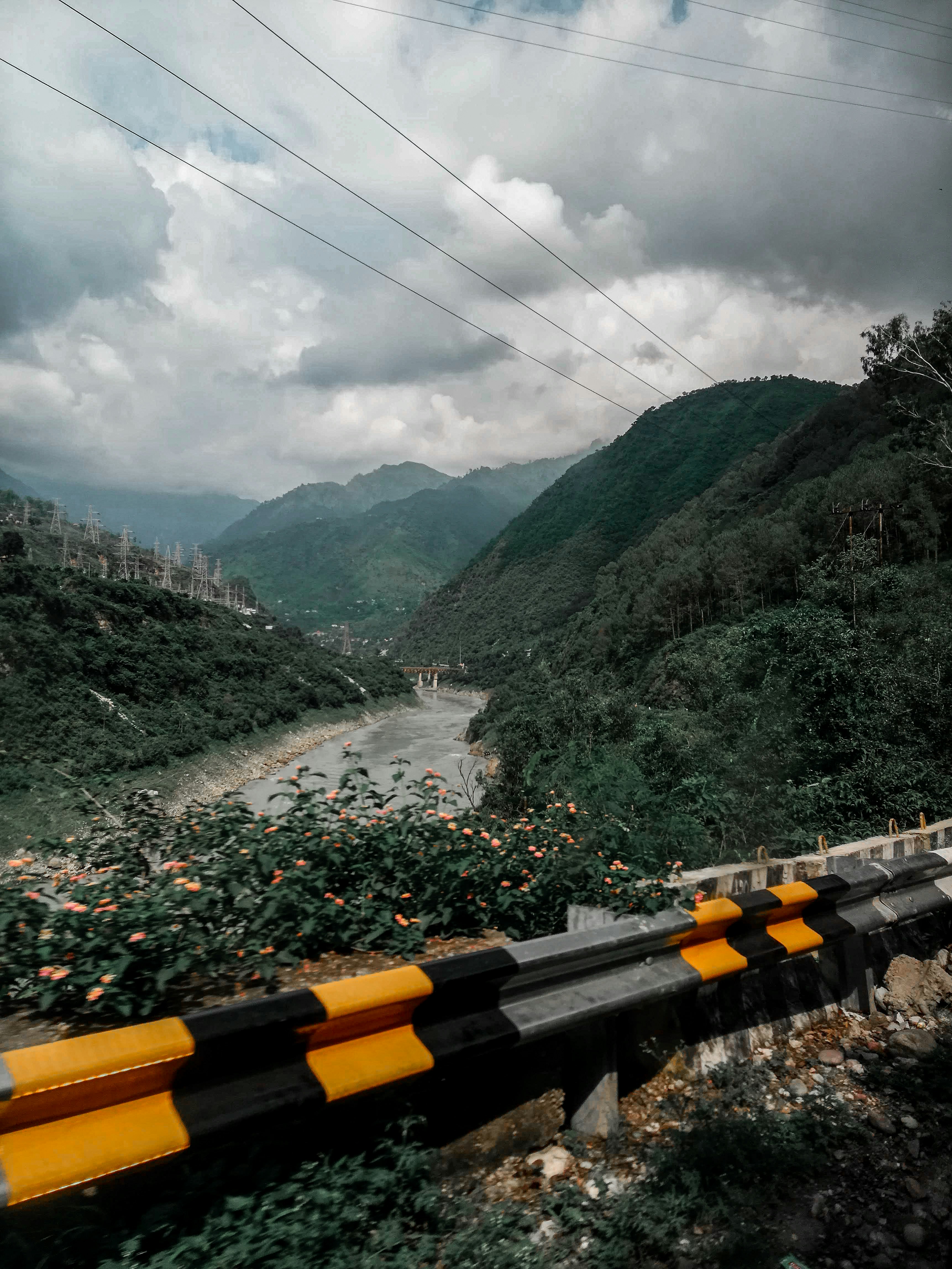 Photograph of a winding river through green mountains, with a yellow-and-black guardrail in the foreground and power lines crossing a cloudy sky.