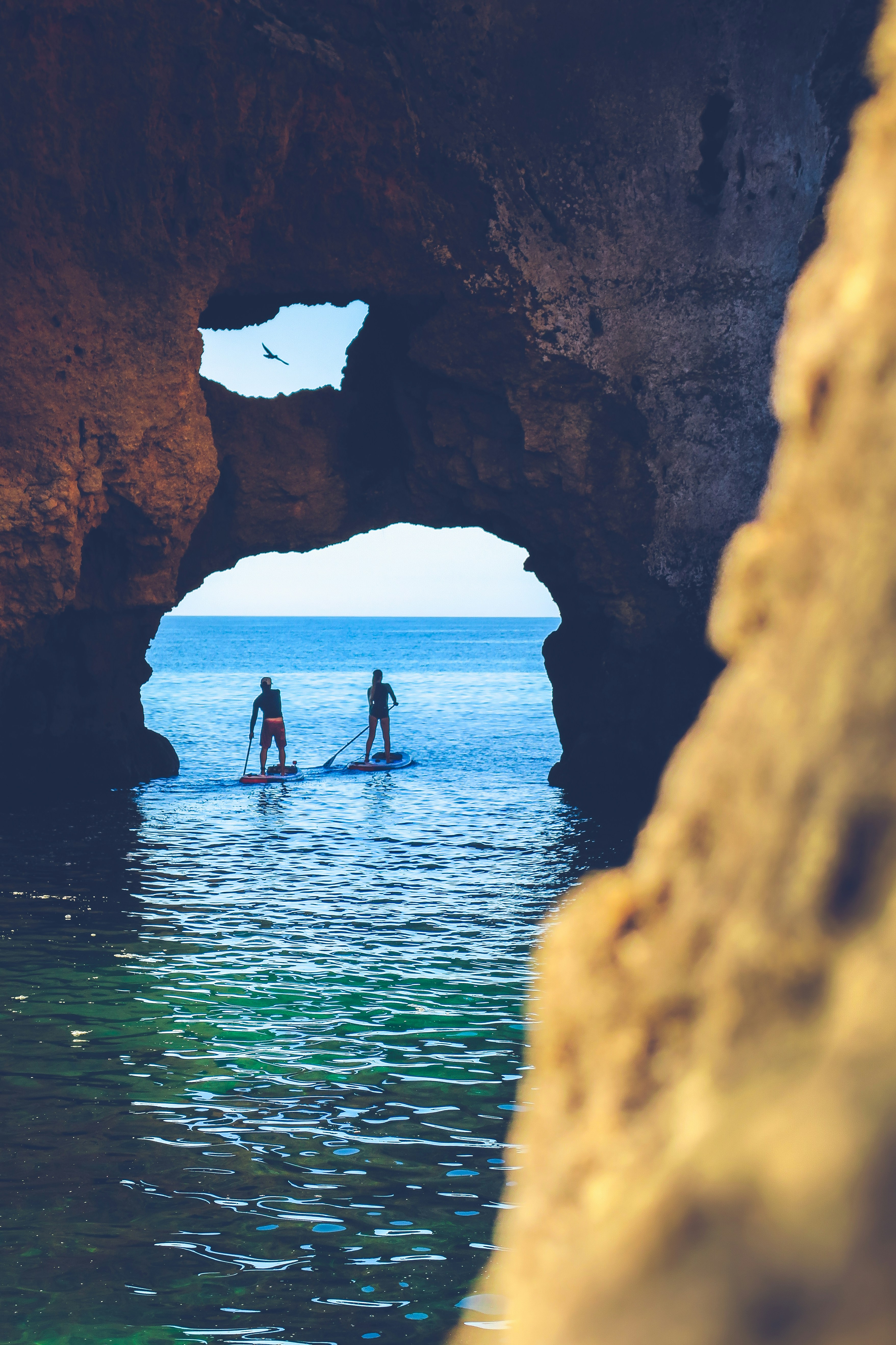 Two paddleboarders glide through a natural rock arch, framed by the vibrant blue sea and a clear sky.