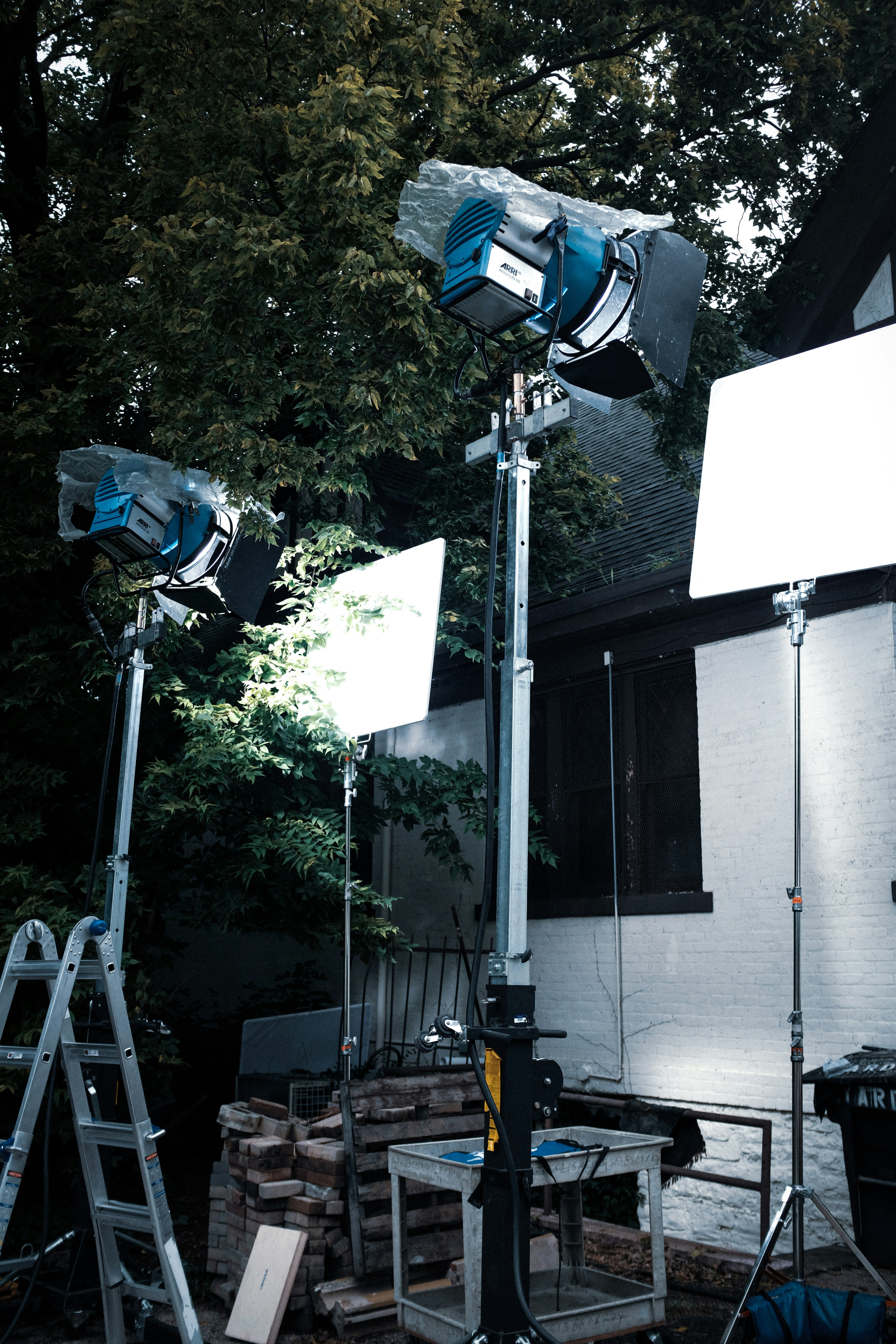 Professional lighting equipment arranged outdoors, showcasing a meticulous setup for a film shoot. Bright lights illuminate the scene amidst lush greenery.