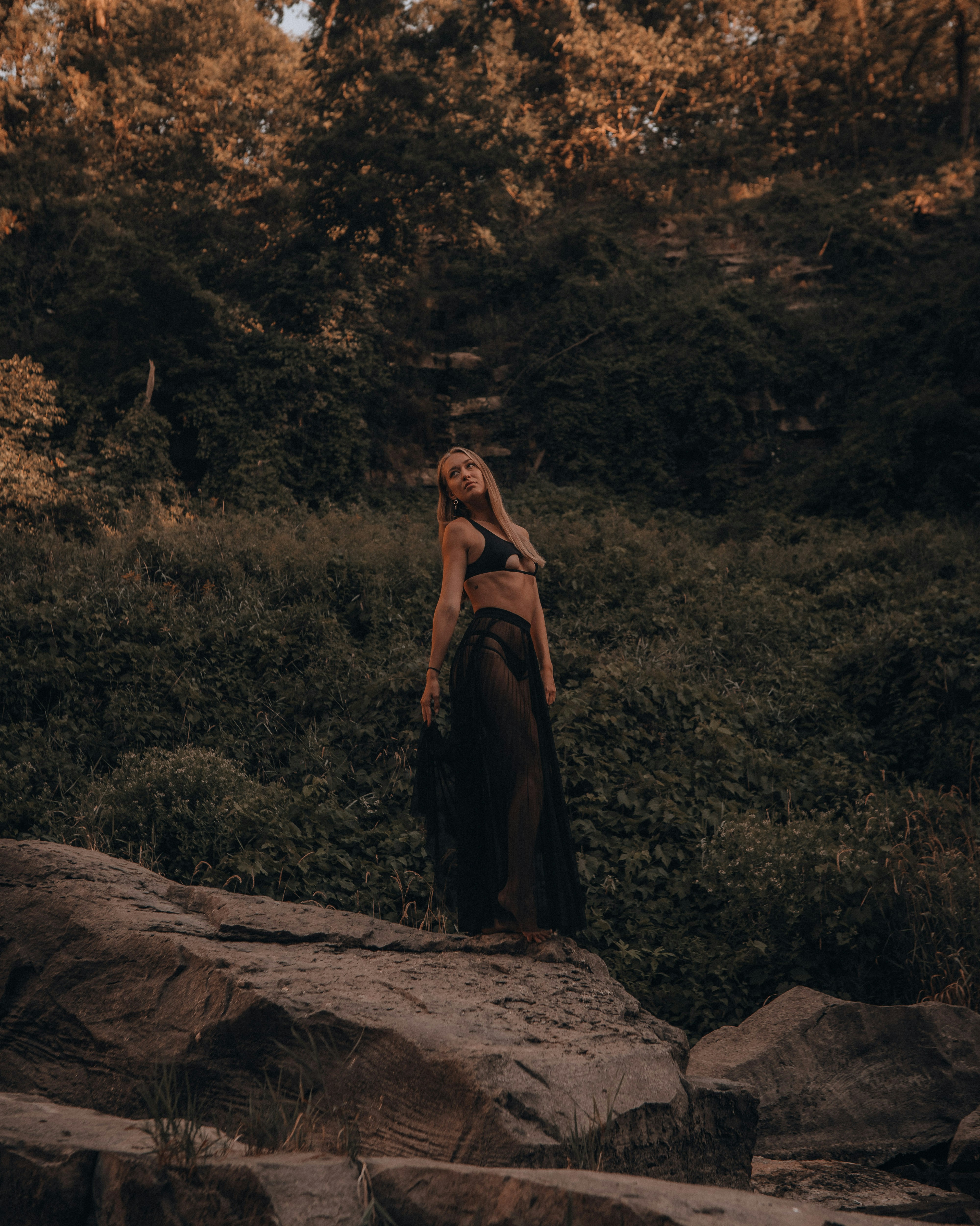 A figure draped in flowing fabric stands gracefully on a rocky outcrop, surrounded by lush greenery and warm, golden light.