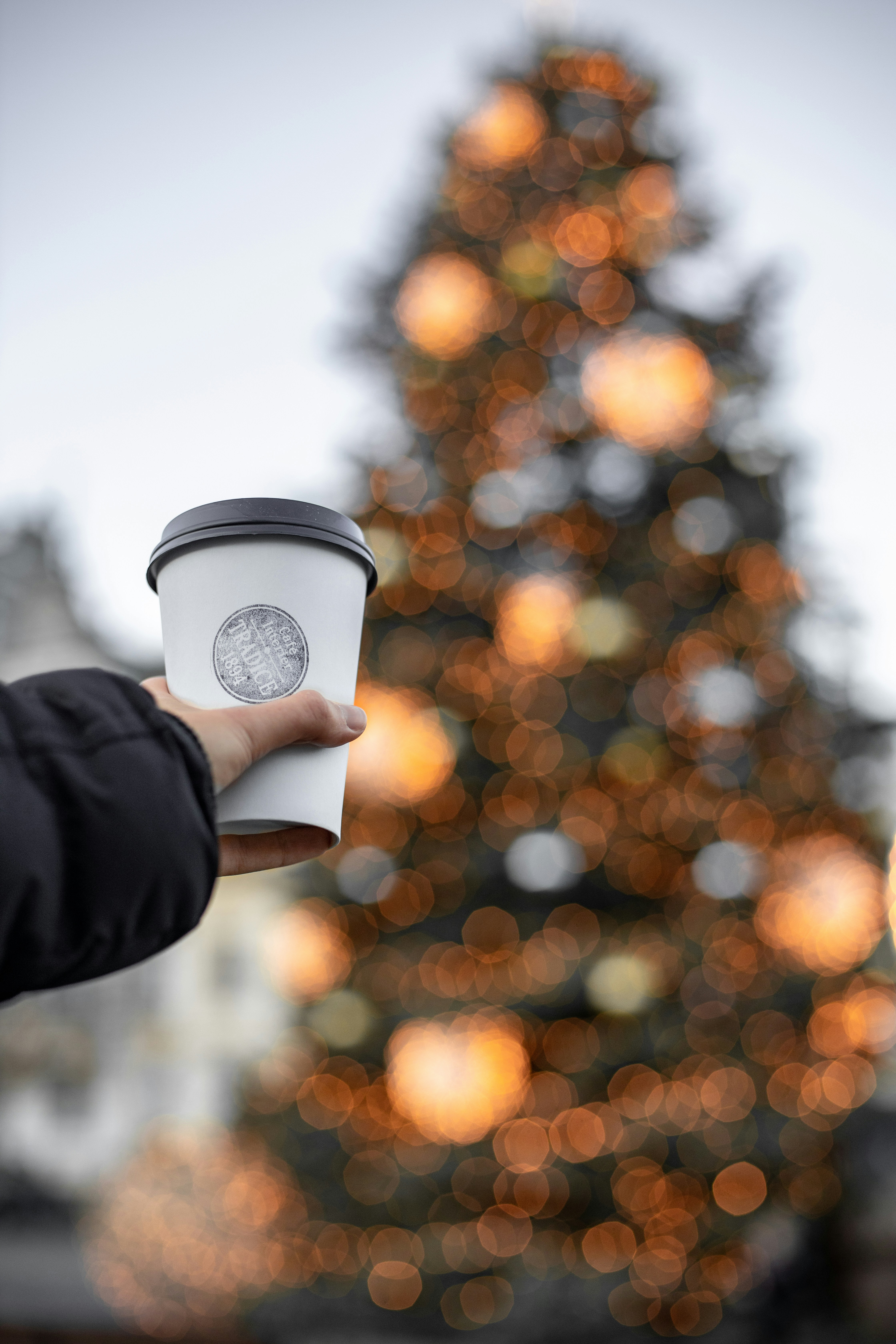 A hand holding a coffee cup in front of a softly blurred Christmas tree adorned with glowing lights.