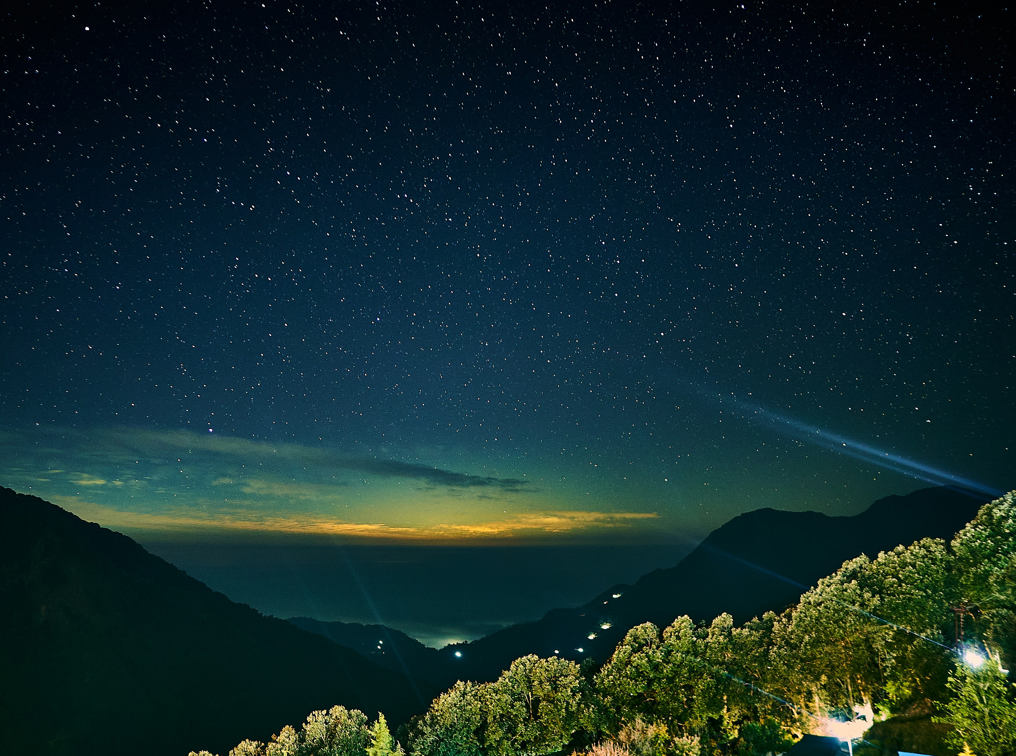 This captivating image showcases a stunning night sky filled with twinkling stars, casting a serene atmosphere over silhouetted mountains. The rich blue and green hues blend seamlessly with the gentle glow of distant lights, creating a harmonious balance of natural and ambient illumination. The composition's depth and expansive star field make it visually striking, inviting viewers to immerse themselves in the tranquil beauty of the scene.