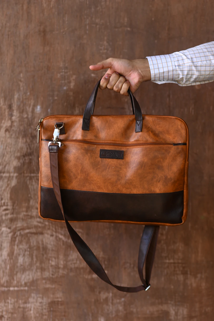 Brown leather briefcase with black trim and shoulder strap being held by a hand against a textured wooden background.
