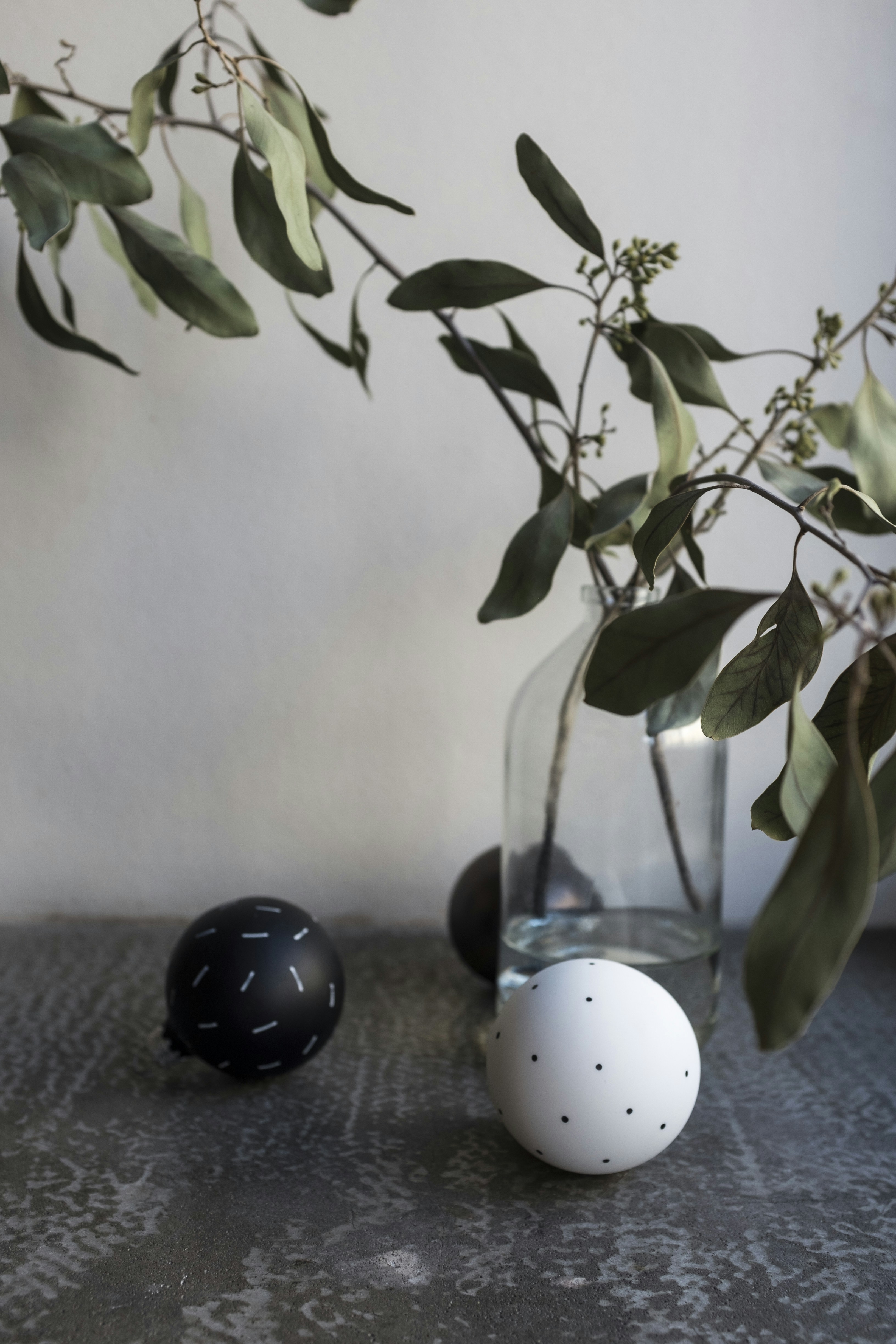 A glass vase filled with greenery sits on a textured surface, accompanied by decorative spheres in black and white. The composition emphasizes simplicity and elegance.
