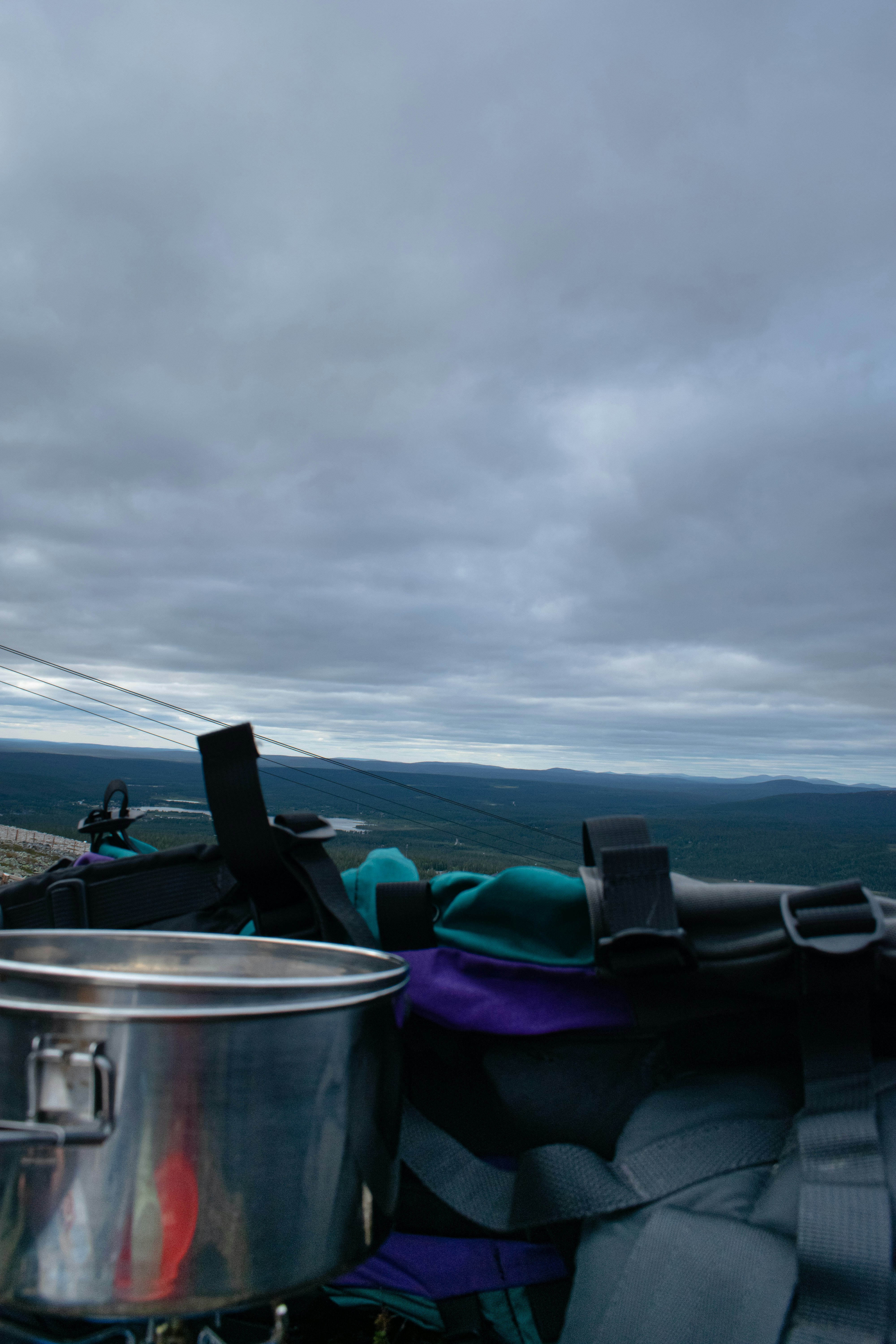 Metal cooking pot with colorful camping gear sits in the foreground on a windswept ridge. A broad, cloudy horizon stretches across the distance.