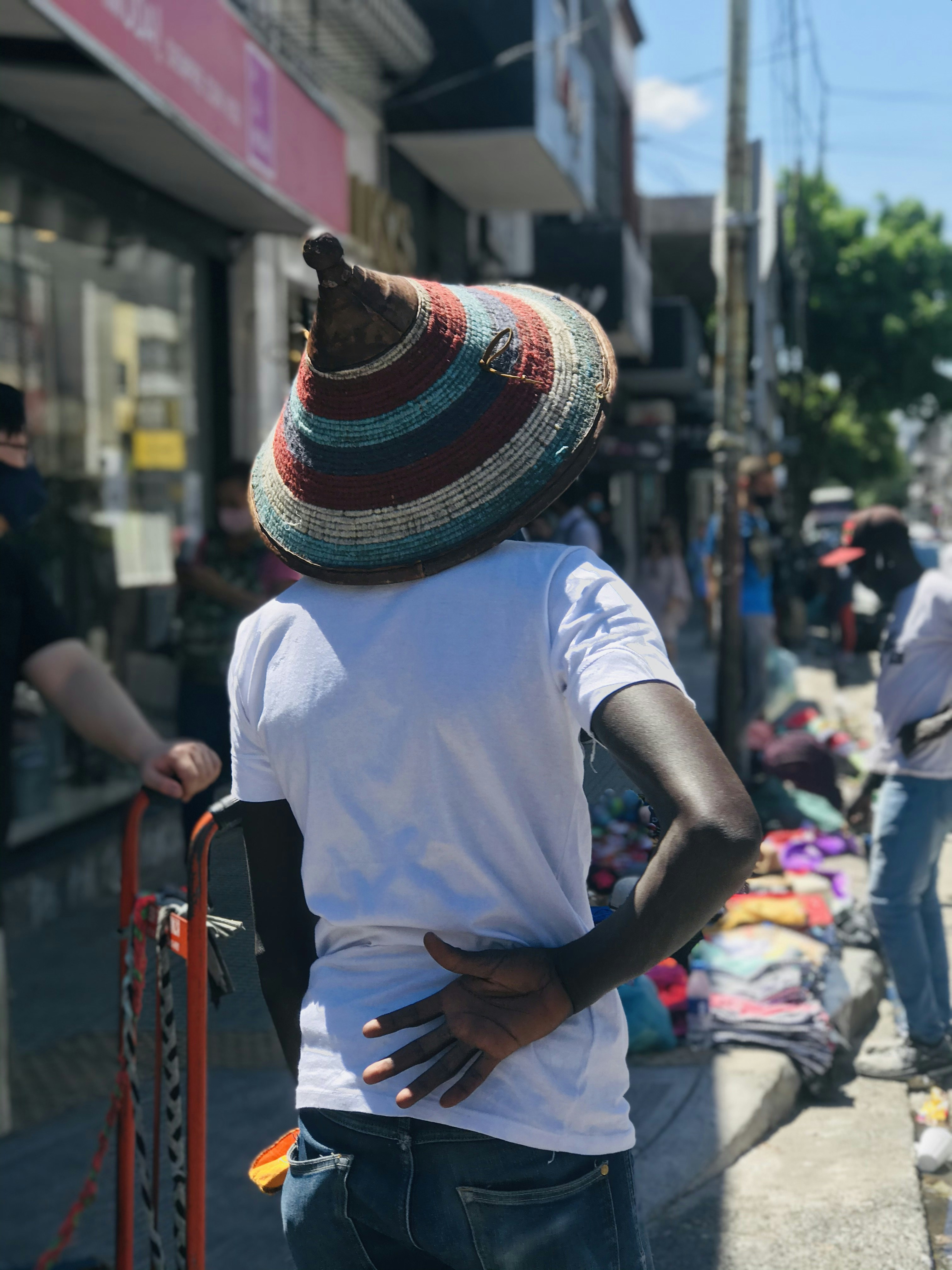 Individual wearing a colorful woven hat stands in a bustling marketplace, observing the vibrant array of textiles laid out on the ground.