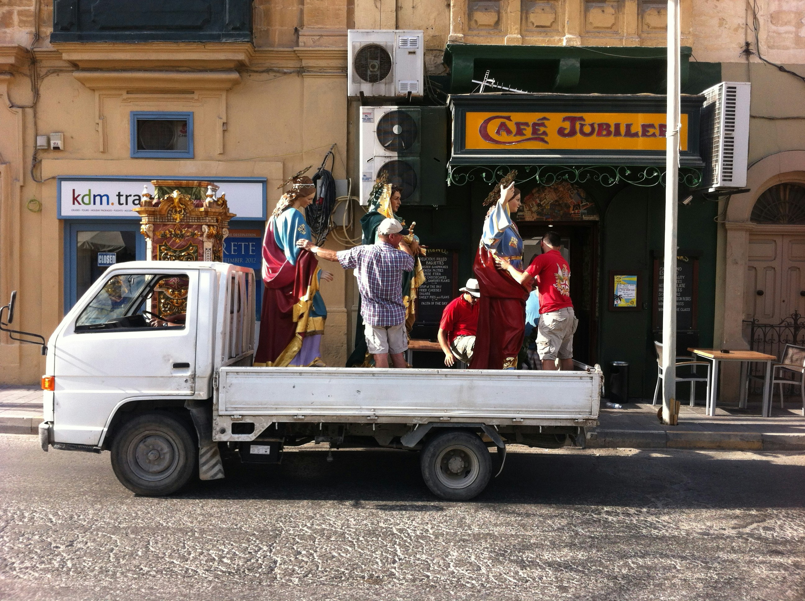Group of people on a small white pickup carrying statues in front of a café; busy urban storefronts with pedestrians and a vehicle in motion.