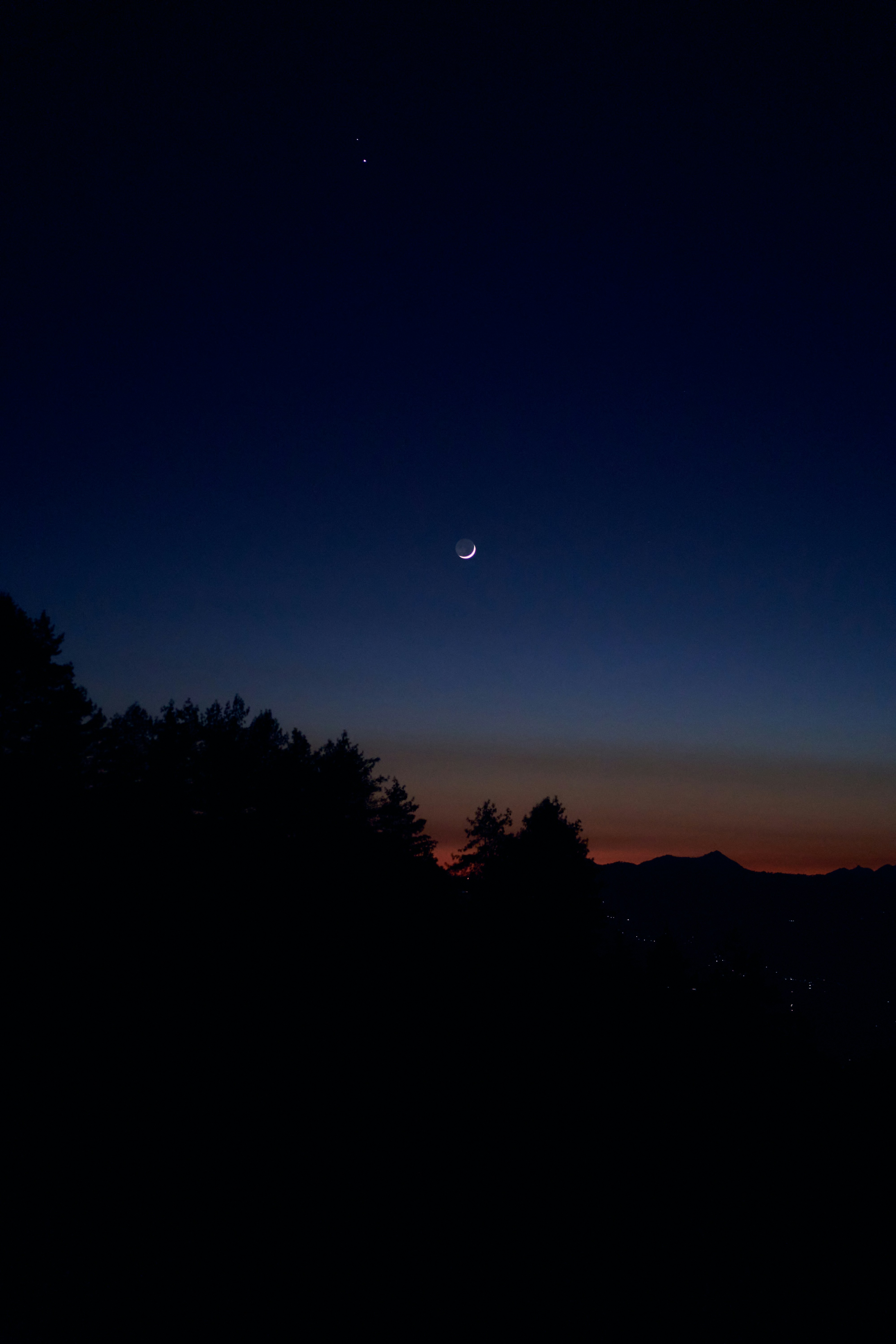 A crescent moon and two distant planets illuminate the twilight sky, framed by silhouetted treetops. The serene transition from day to night captures the essence of cosmic beauty.