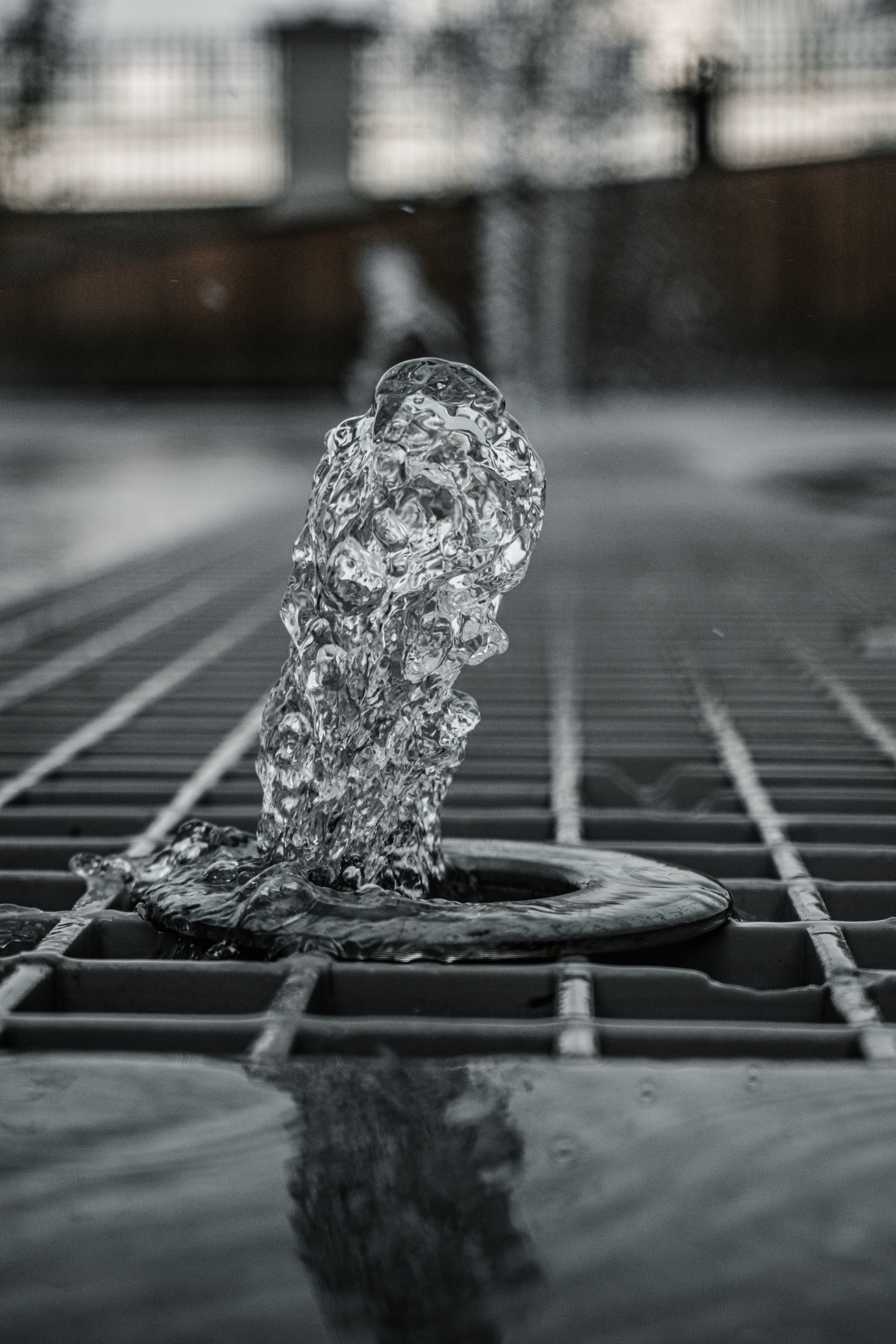 Water fountain spray emerging from a circular drain cover, captured in monochrome. The rippling water reflects the surrounding environment.
