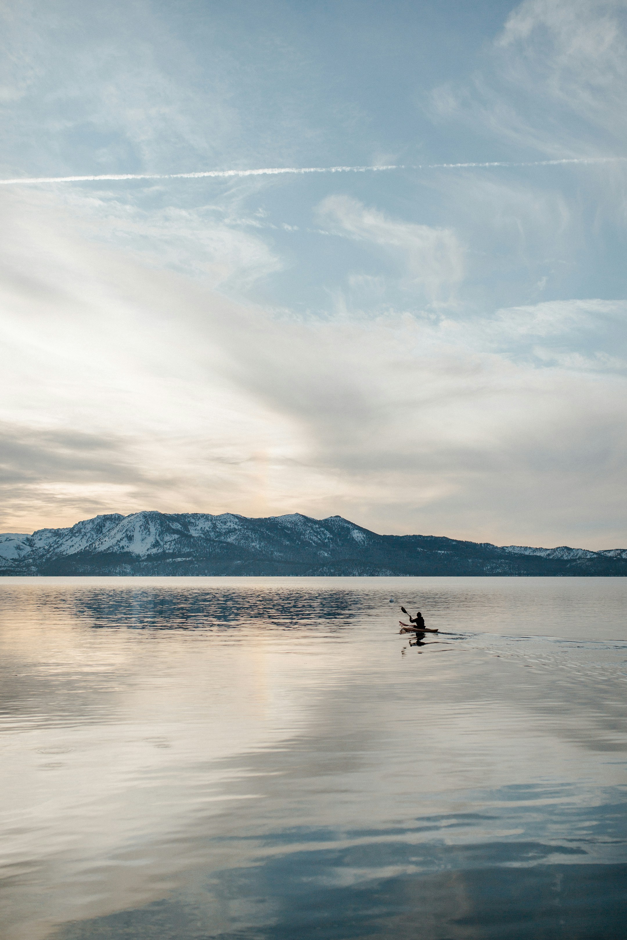 A solitary kayaker glides across a tranquil lake, framed by distant snow-capped mountains under a soft, cloudy sky.