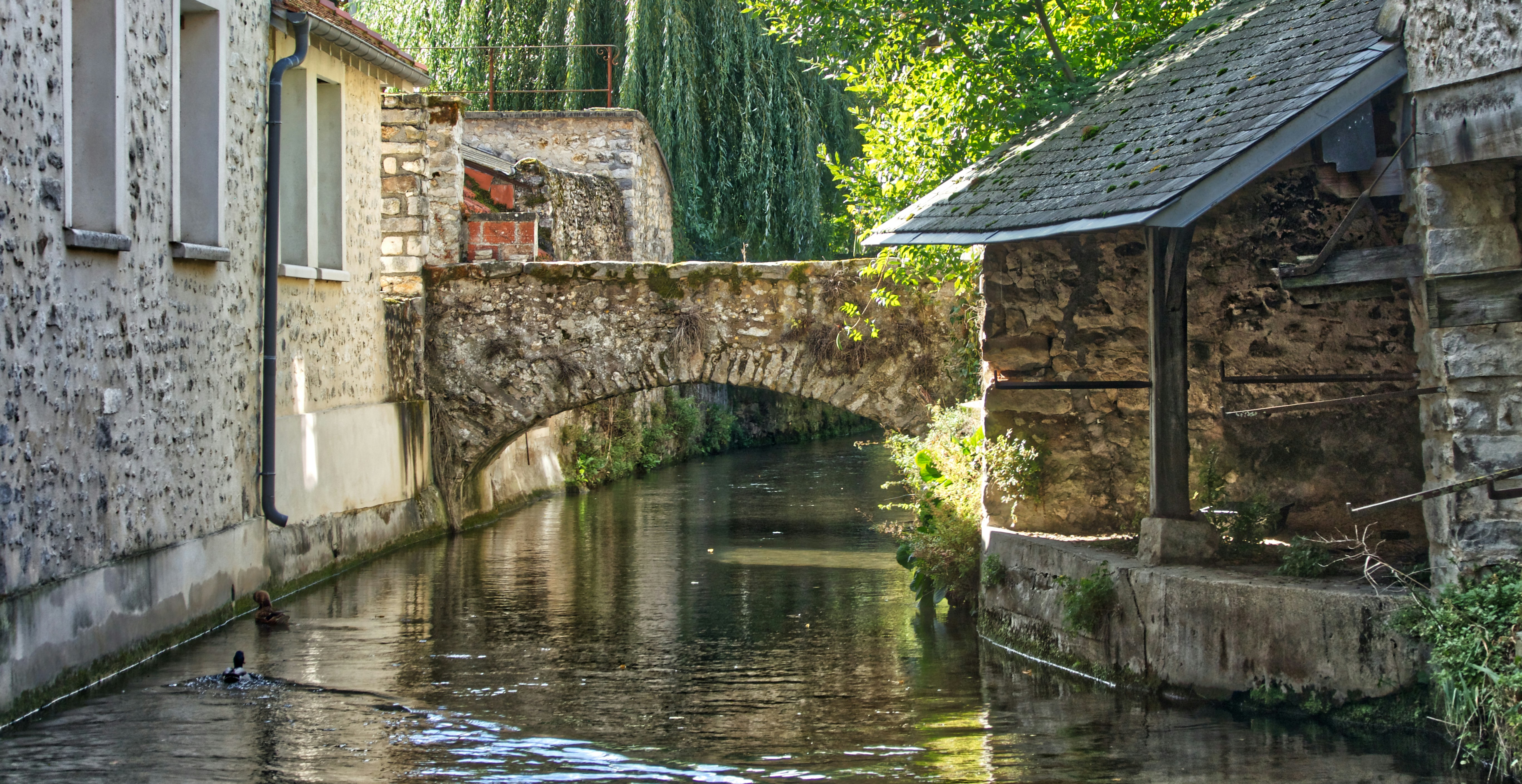 A serene canal scene featuring a quaint stone bridge and rustic buildings, surrounded by lush greenery. The calm water reflects the tranquil atmosphere.