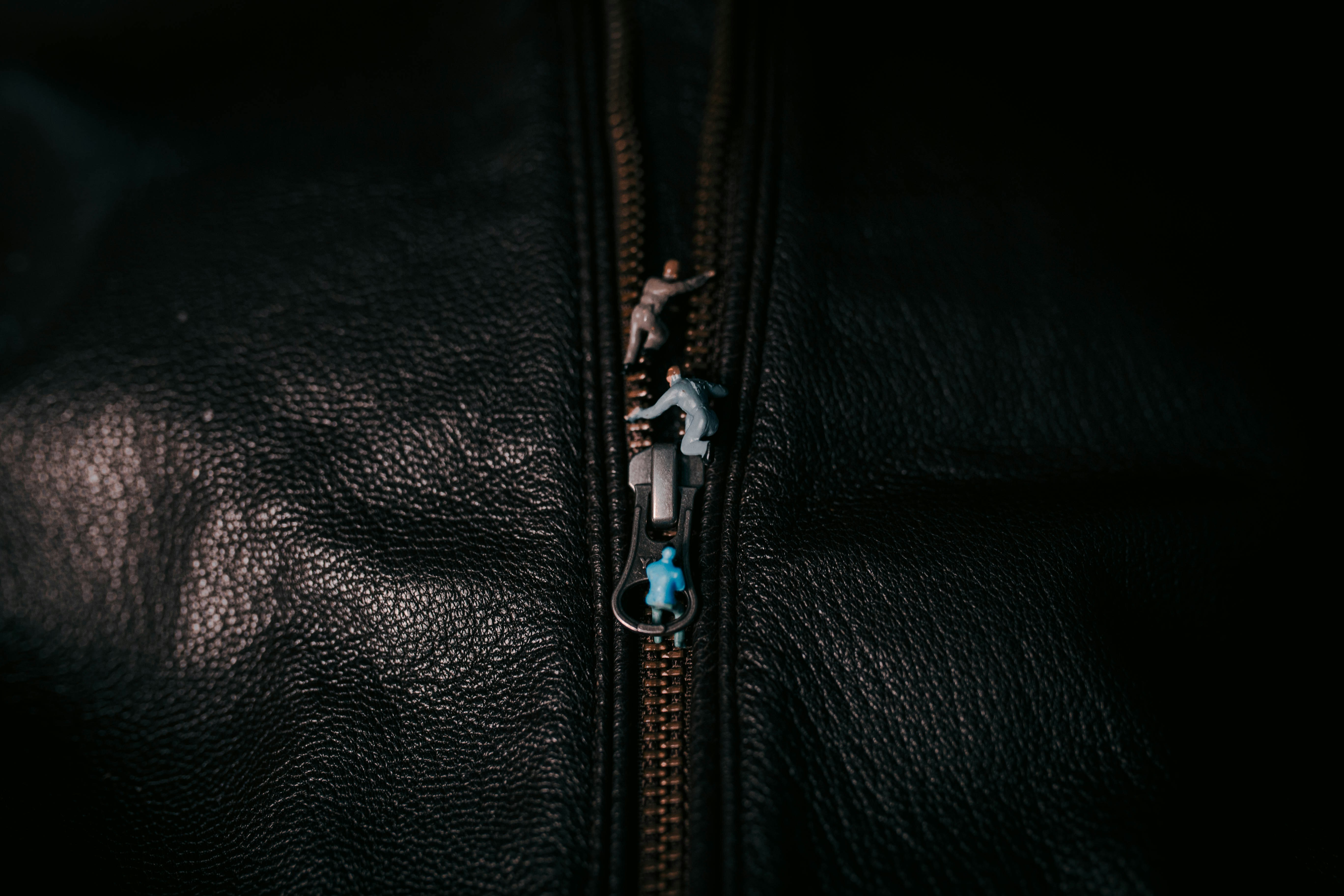 Two tiny figurines scaling a zipper on a black leather surface, creating a playful juxtaposition of scale and texture.