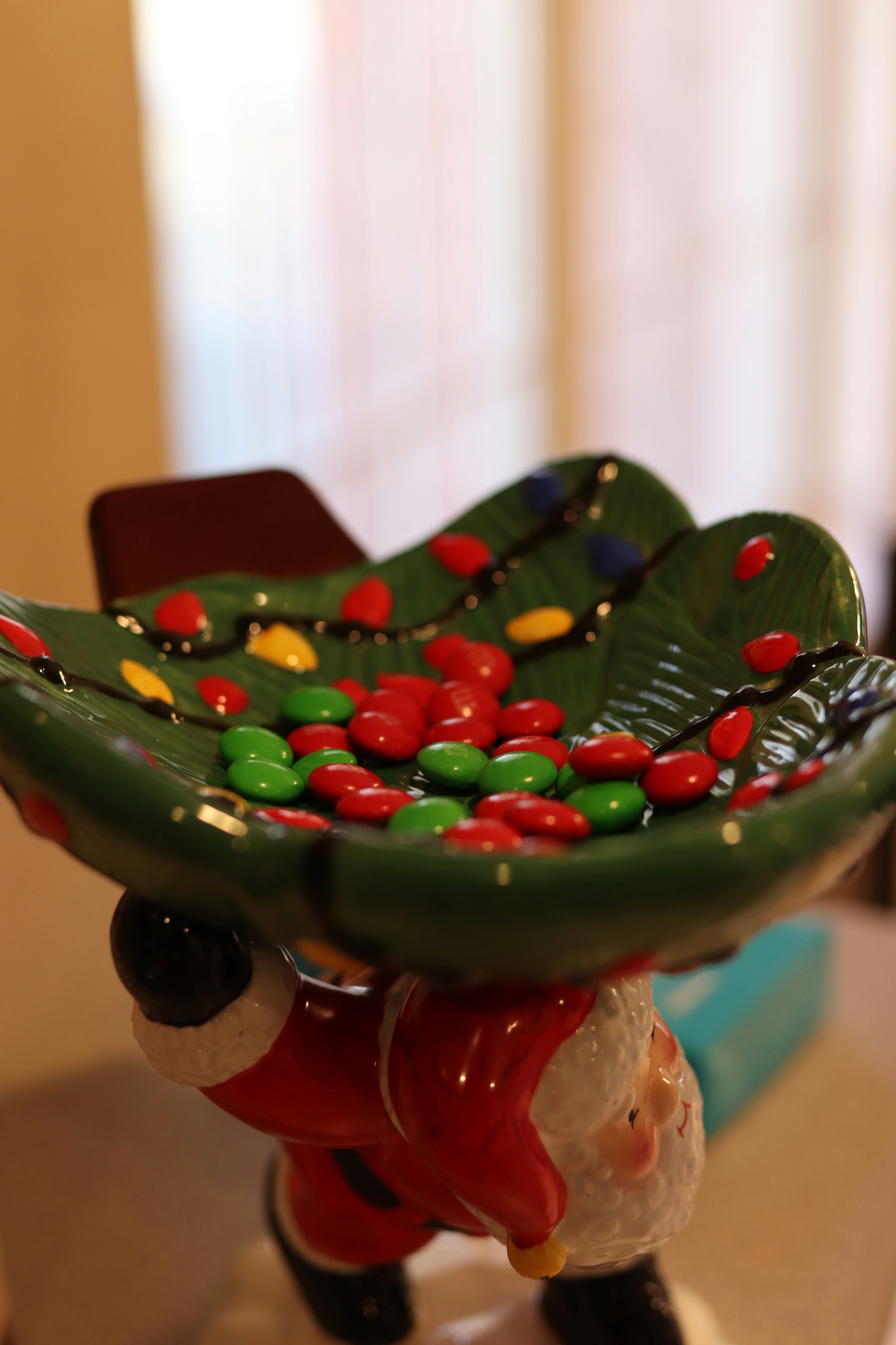 A whimsical green bowl shaped like a leaf, filled with colorful candies, rests atop a Santa figurine. The background features soft, blurred light, enhancing the festive atmosphere.
