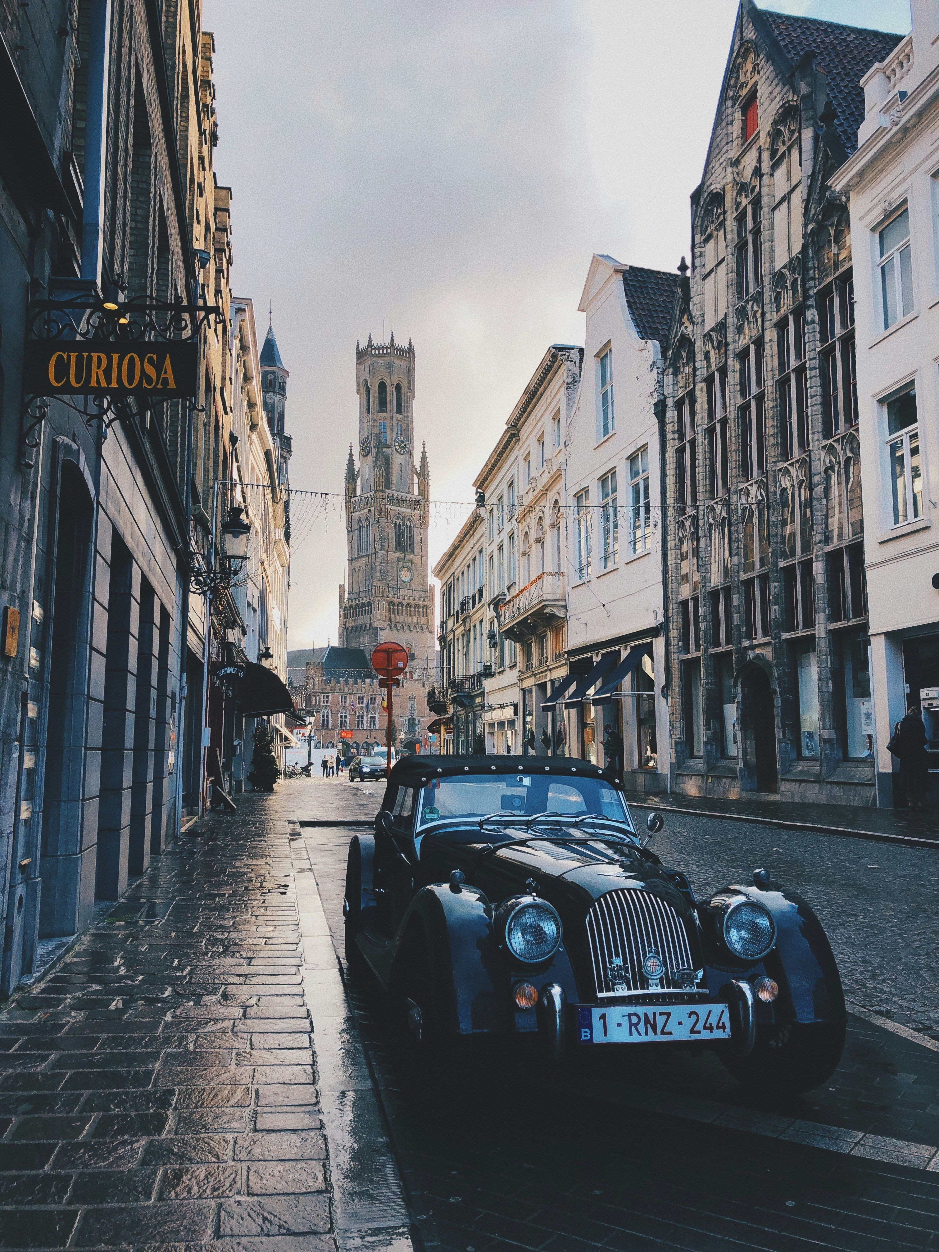 A vintage black car parked on a cobblestone street, framed by historic buildings and a towering clock tower in the background.