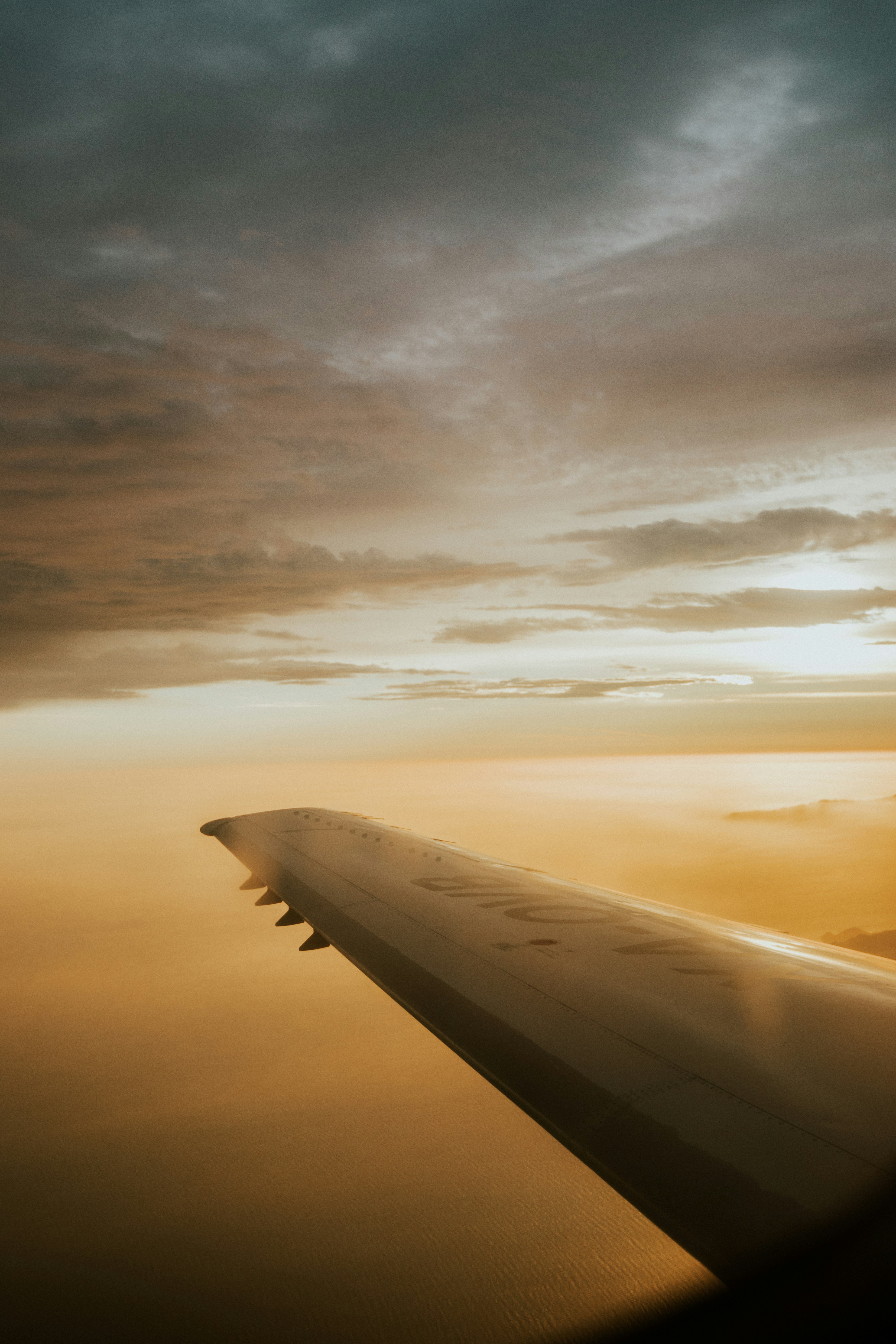 Airplane wing extending over a vast horizon, bathed in warm golden light during sunset. The clouds create a dramatic backdrop.