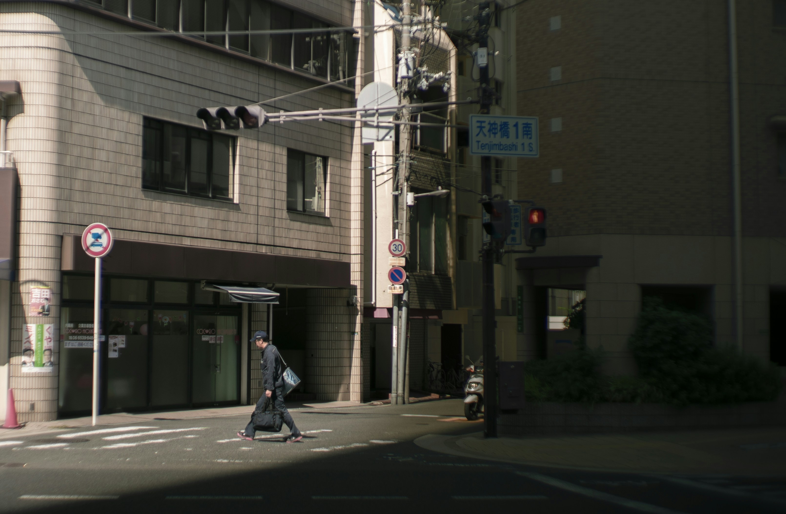 A lone figure crosses a quiet street in a modern city, framed by buildings and traffic signals. The scene captures the essence of urban life.