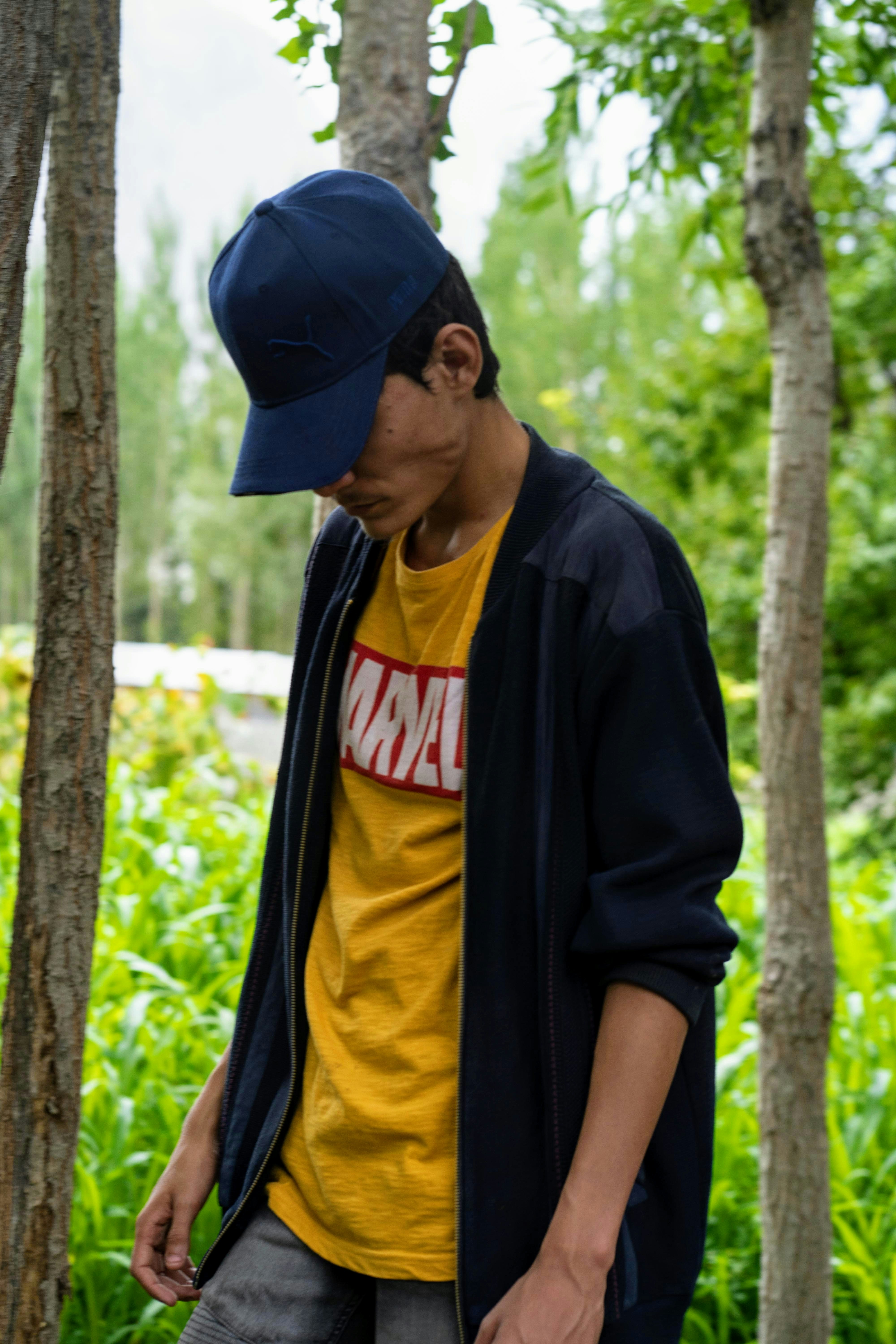 Young man in a yellow Marvel shirt and black jacket, standing amidst lush greenery, lost in thought.