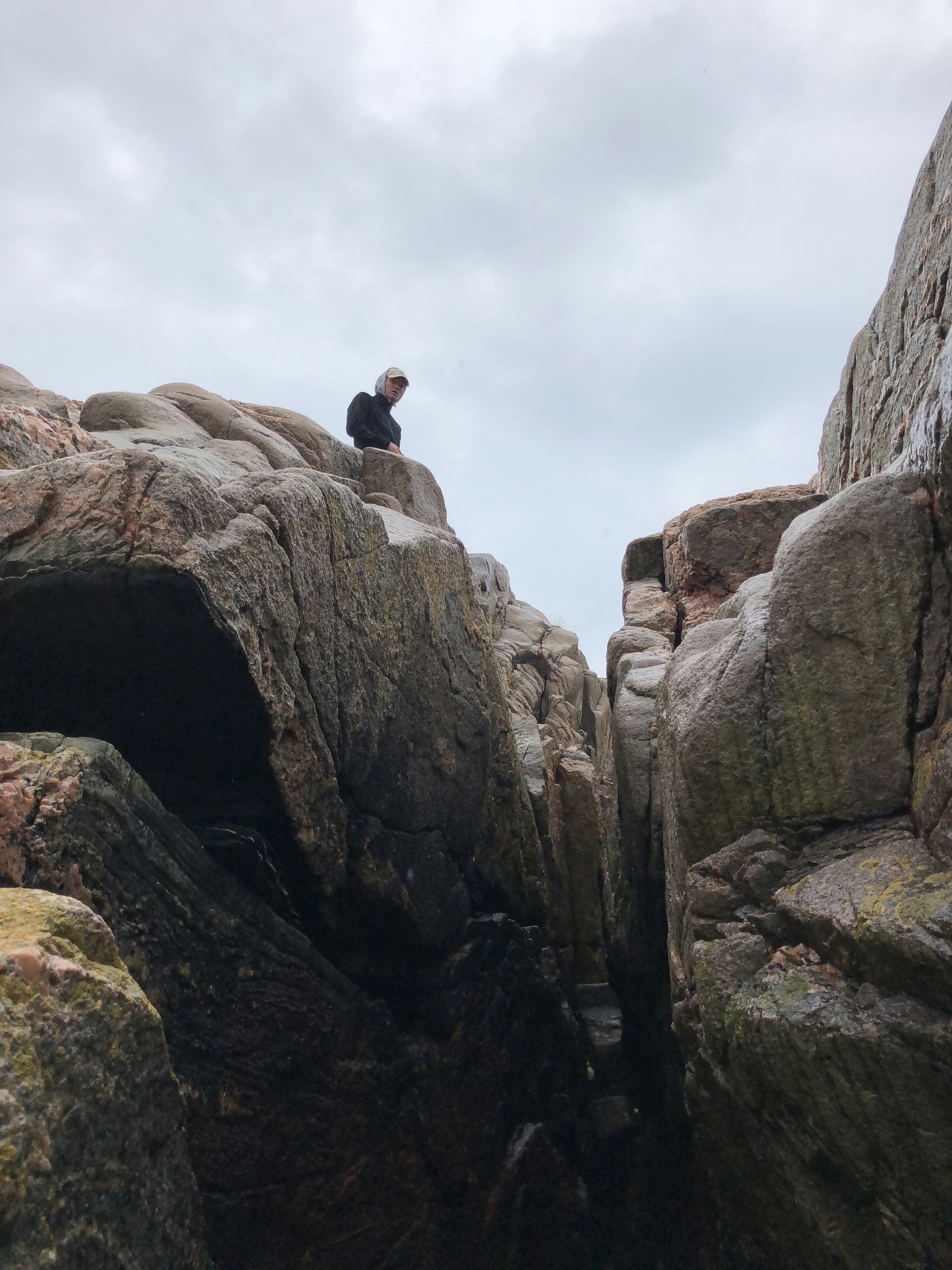 A figure stands atop rugged rocks, framed by a narrow crevice, under a cloudy sky. The scene evokes a sense of solitude and adventure.